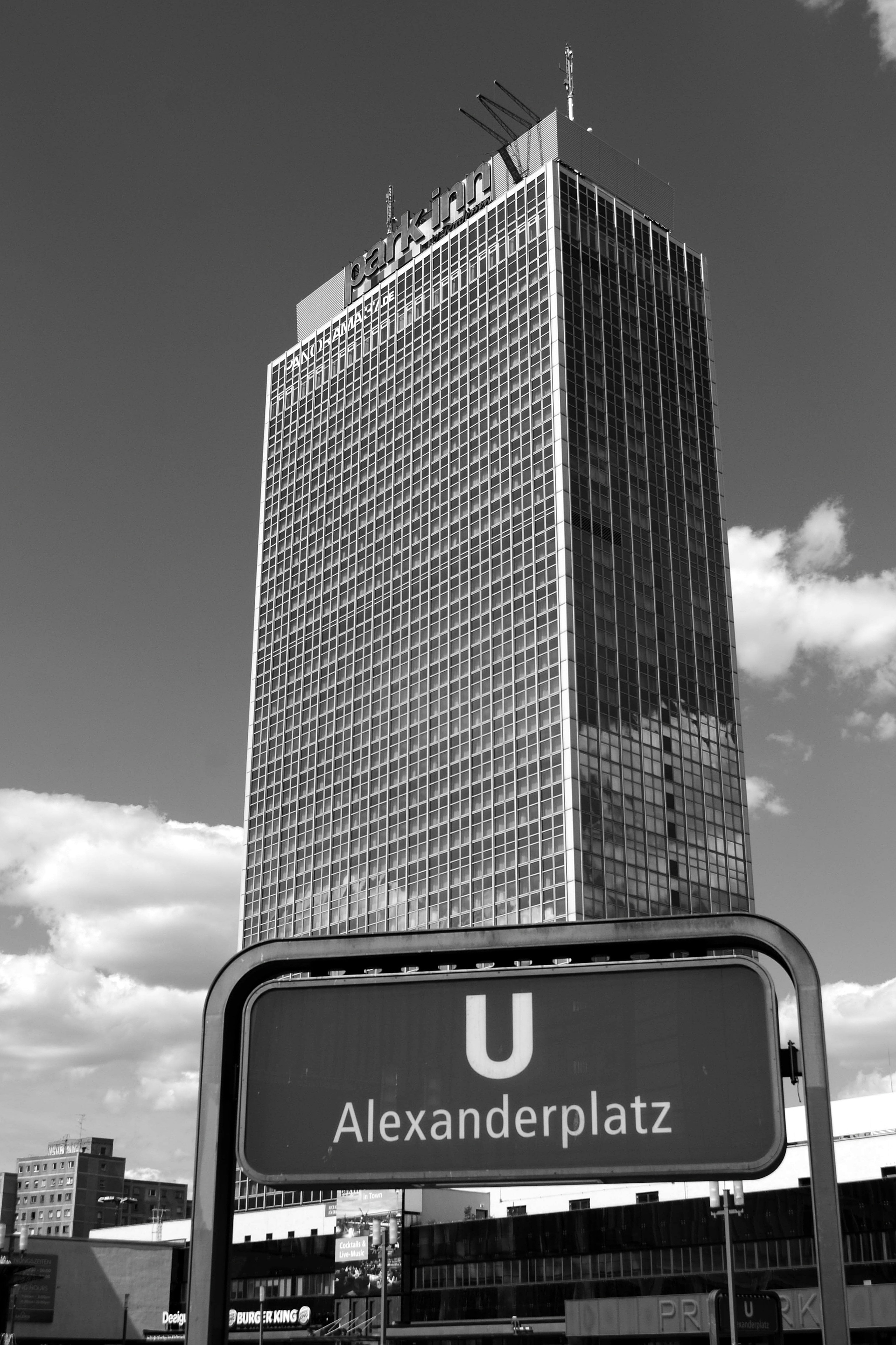 Tall modern building looms over the iconic Alexanderplatz sign, framed by a dramatic sky. A striking contrast of architecture and urban life.