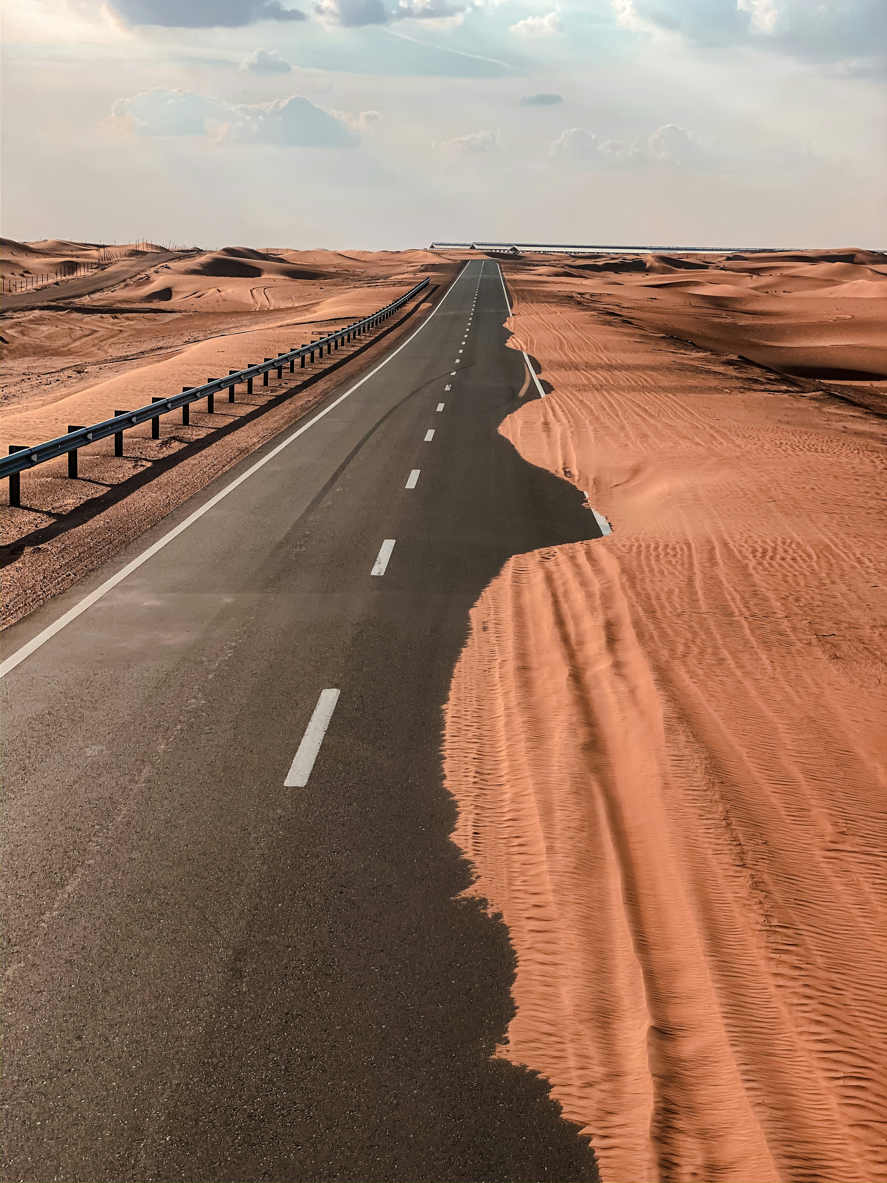 A winding road cuts through expansive desert dunes under a cloudy sky. The contrast between the asphalt and the sandy terrain creates a striking visual narrative.