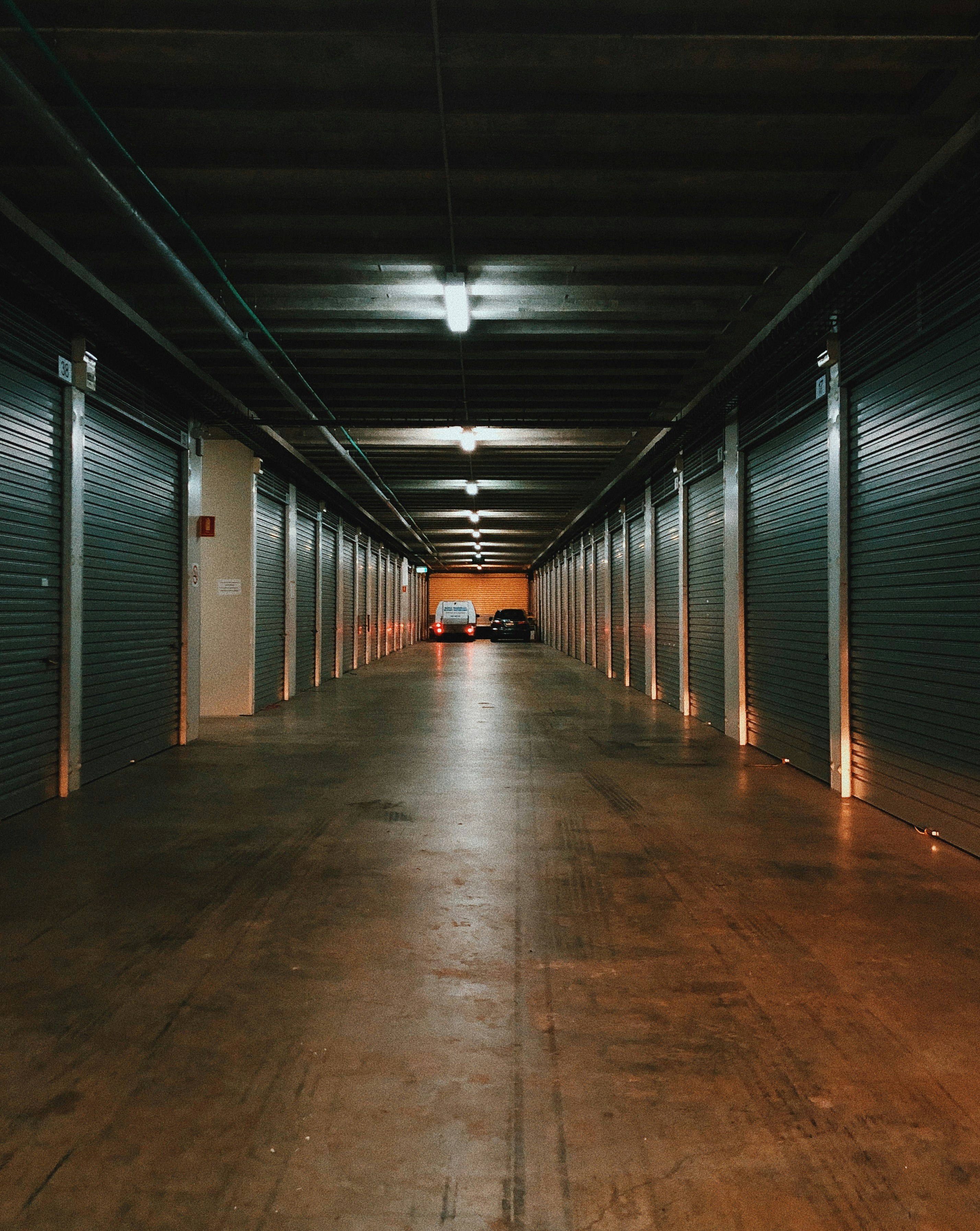 Dimly lit parking garage with rows of closed storage units and two parked vehicles at the end, creating a sense of depth and perspective.