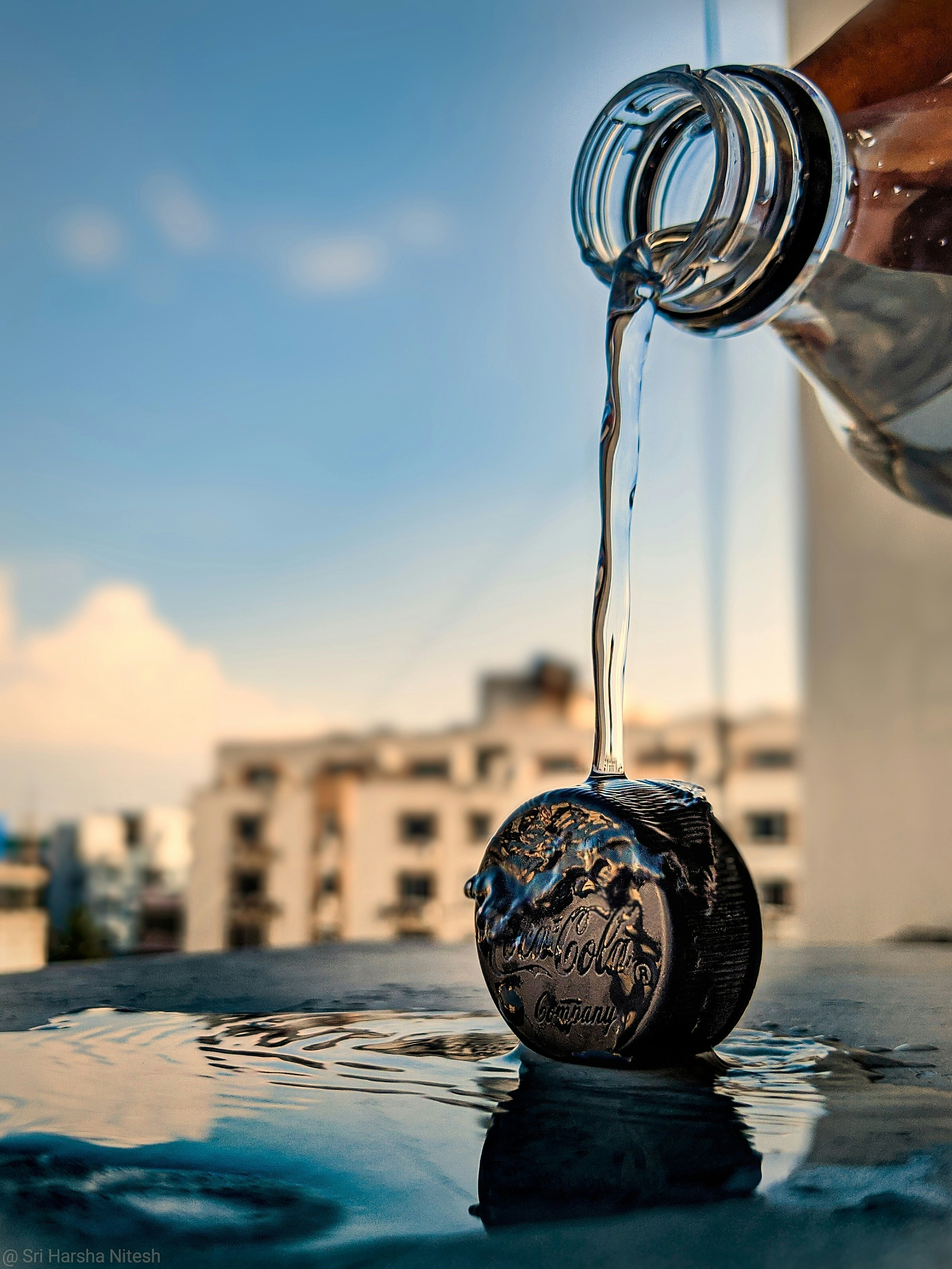 Water cascading from a bottle onto a metallic cap, creating ripples in a puddle. The background features blurred urban architecture.