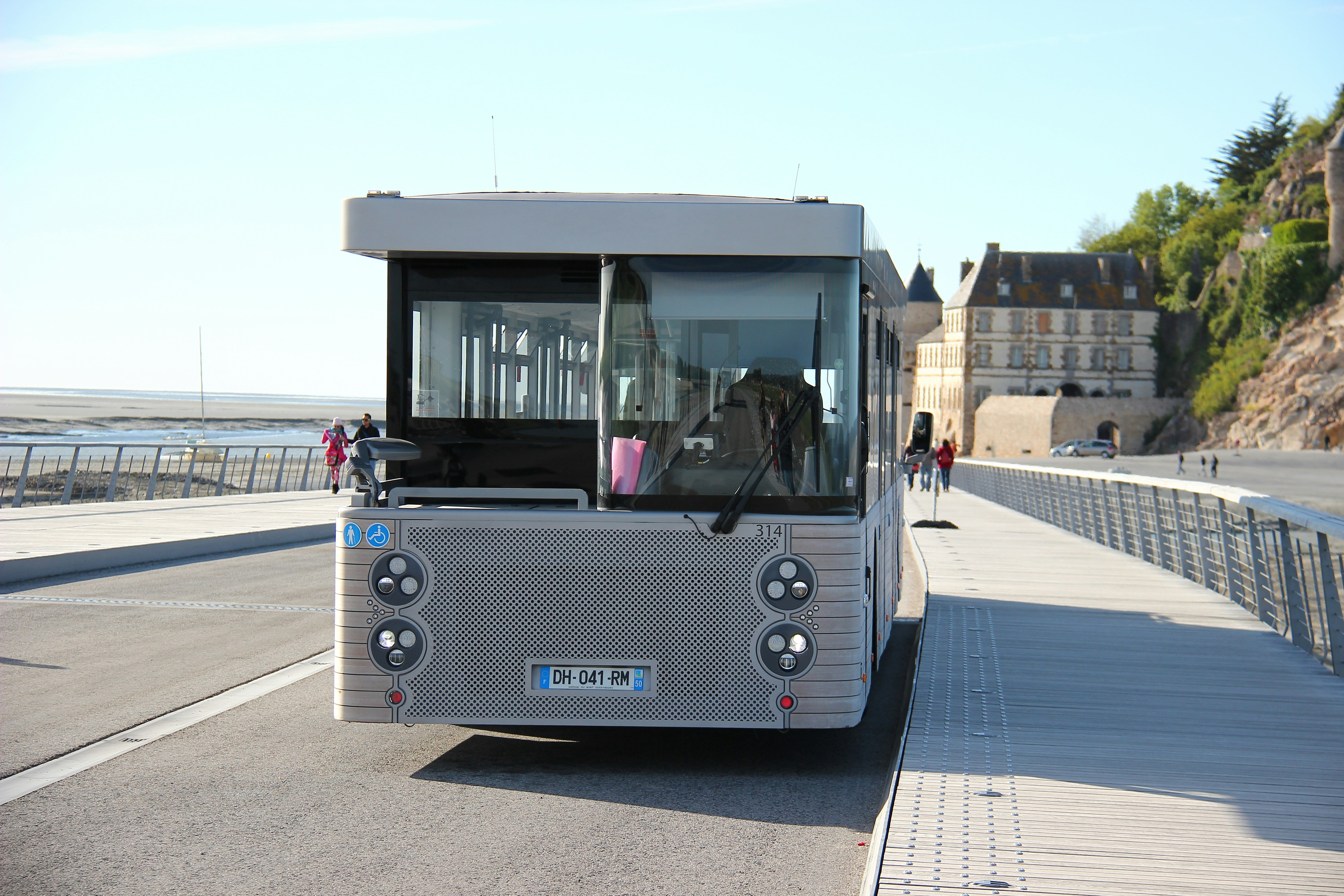 A sleek, modern bus stands on a boardwalk with historical buildings in the background, showcasing a blend of contemporary design and classic architecture.