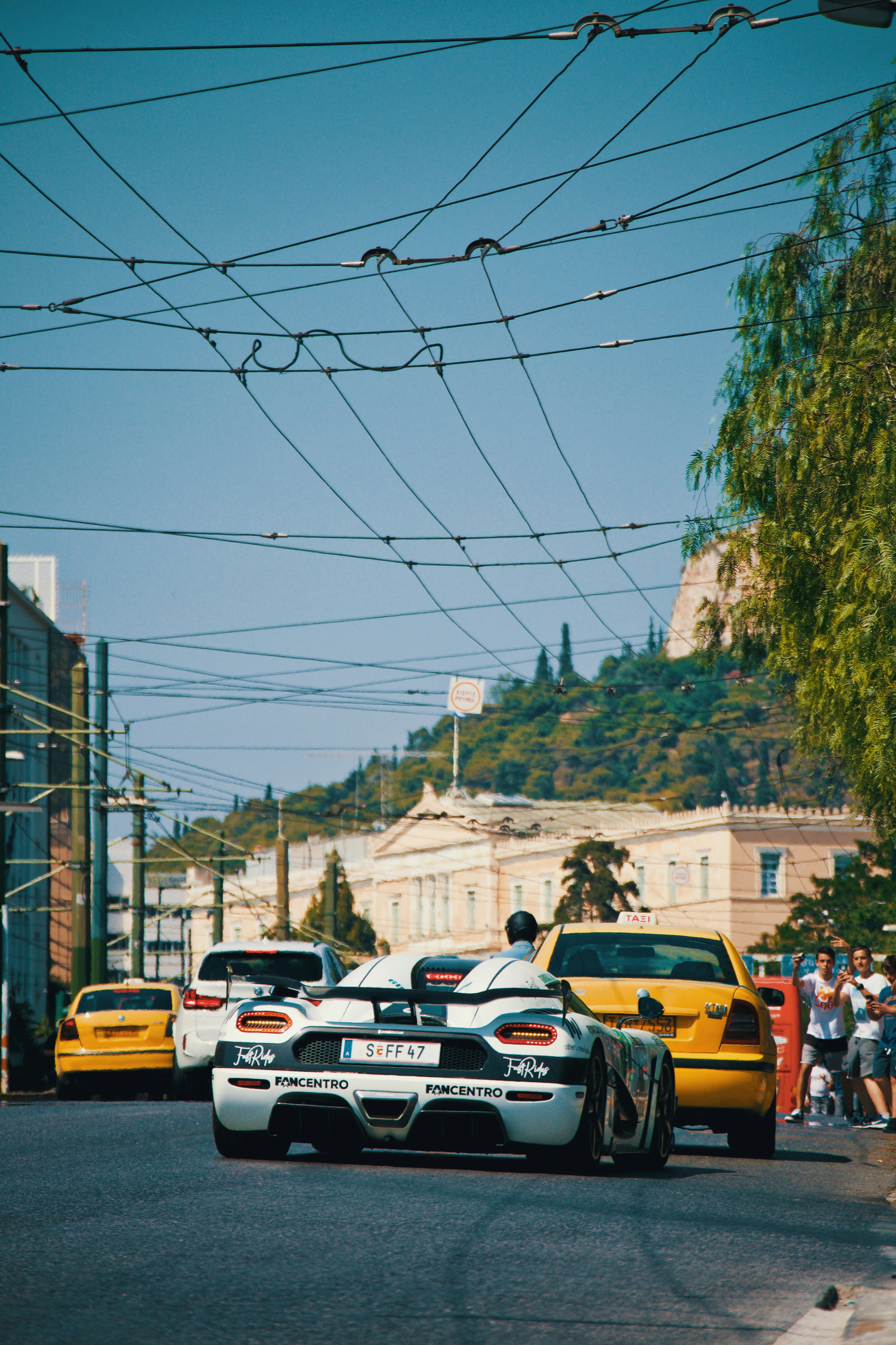 A sleek white sports car maneuvers through a bustling city street, flanked by taxis and onlookers. Power lines crisscross the sky, framing the scene.