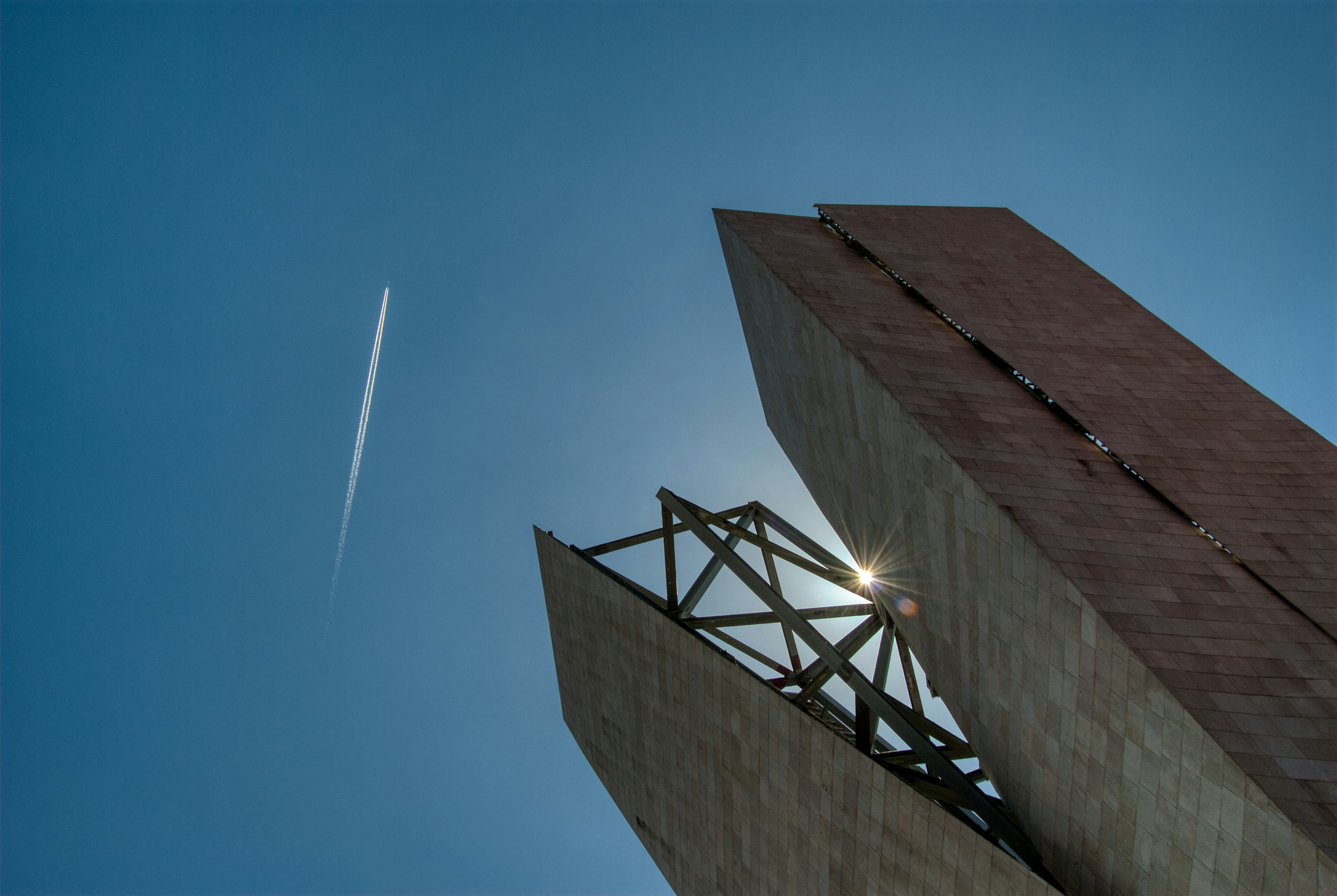 Architectural structure with sunlit angles against a clear blue sky, intersected by a jet's contrail.