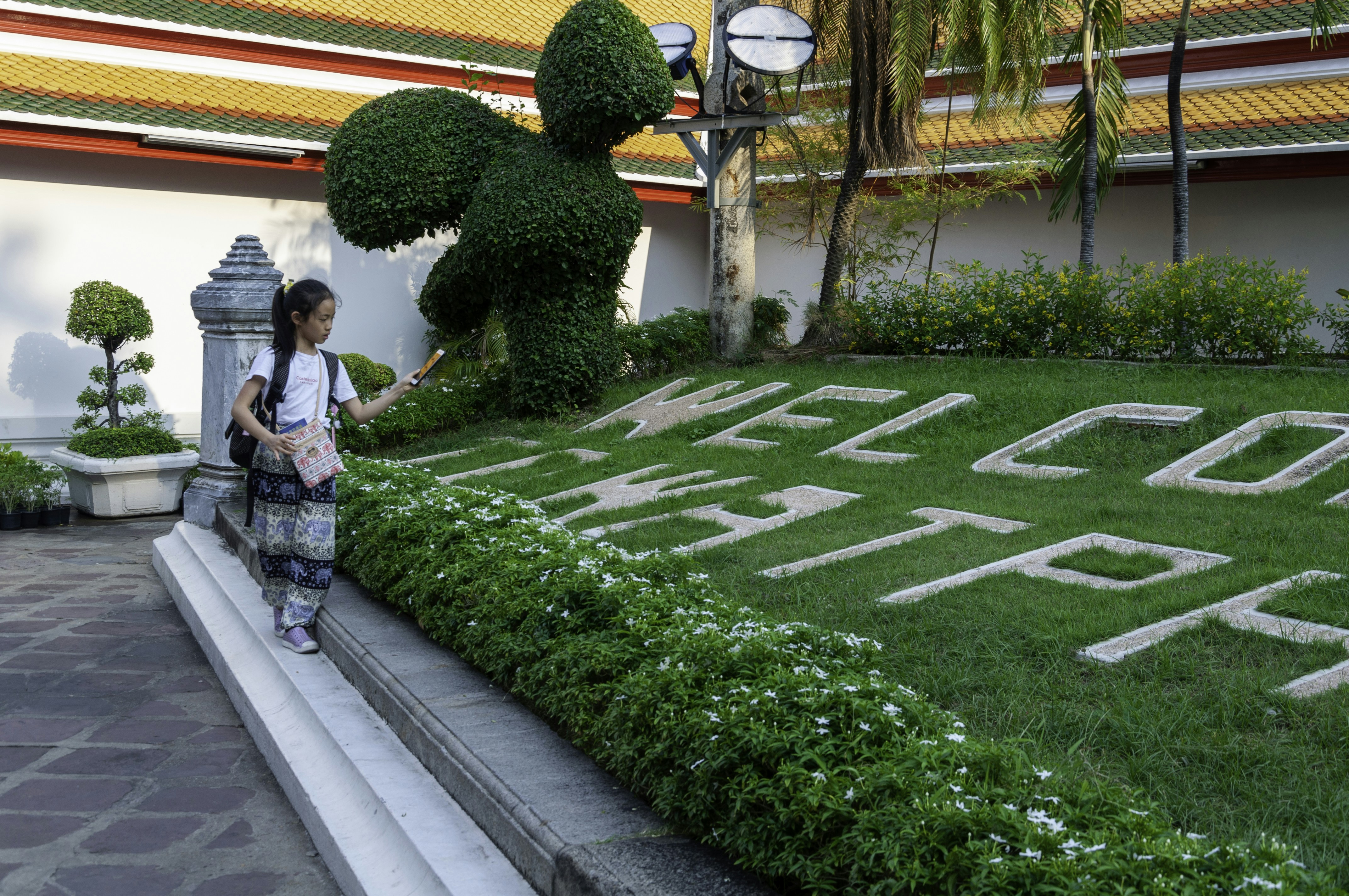 Person photographing whimsical topiary near a garden with 'WELCOME' spelled in plants.