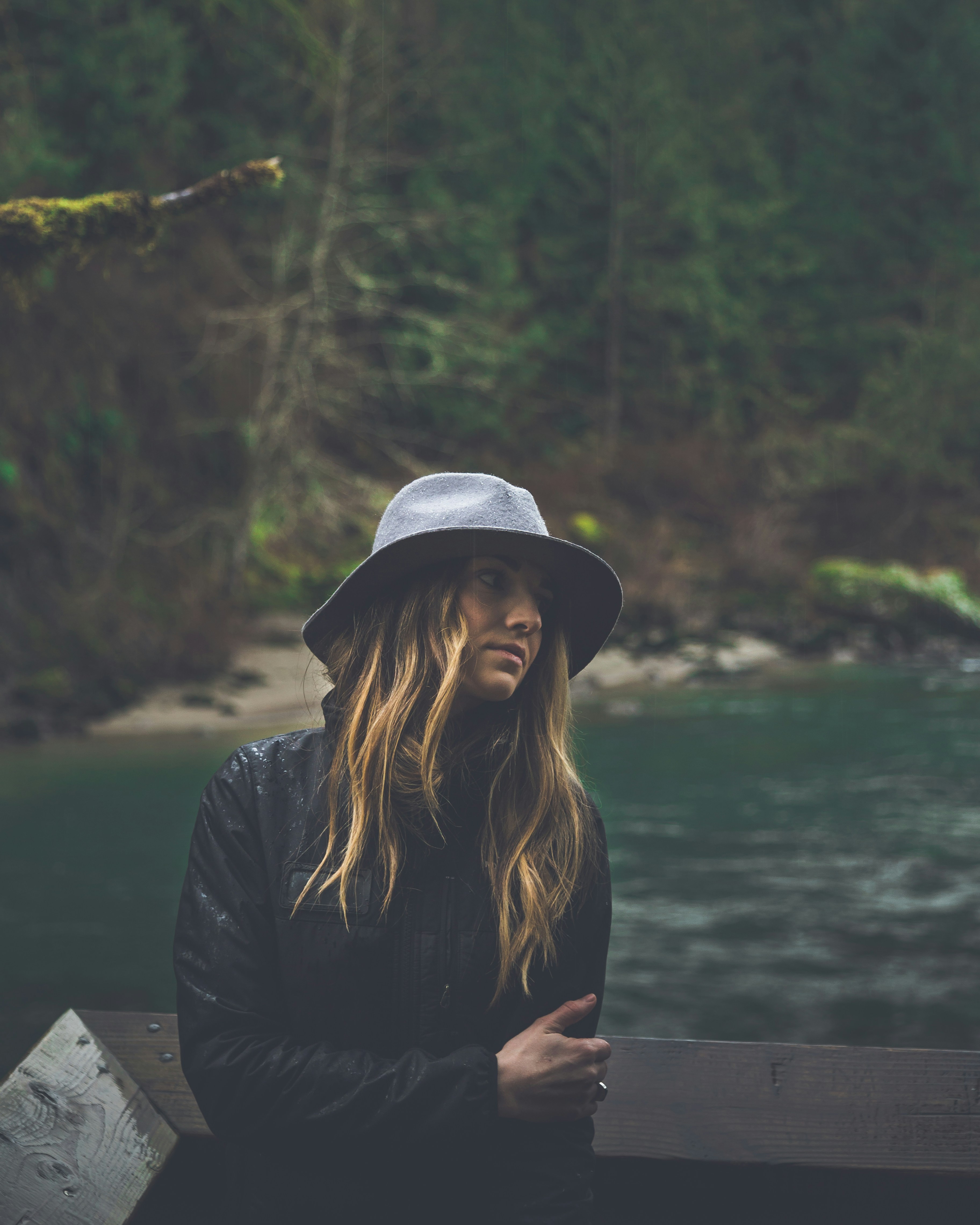 A contemplative figure stands by a tranquil river, adorned in a dark coat and wide-brimmed hat, surrounded by lush greenery. The scene evokes a serene connection with nature.