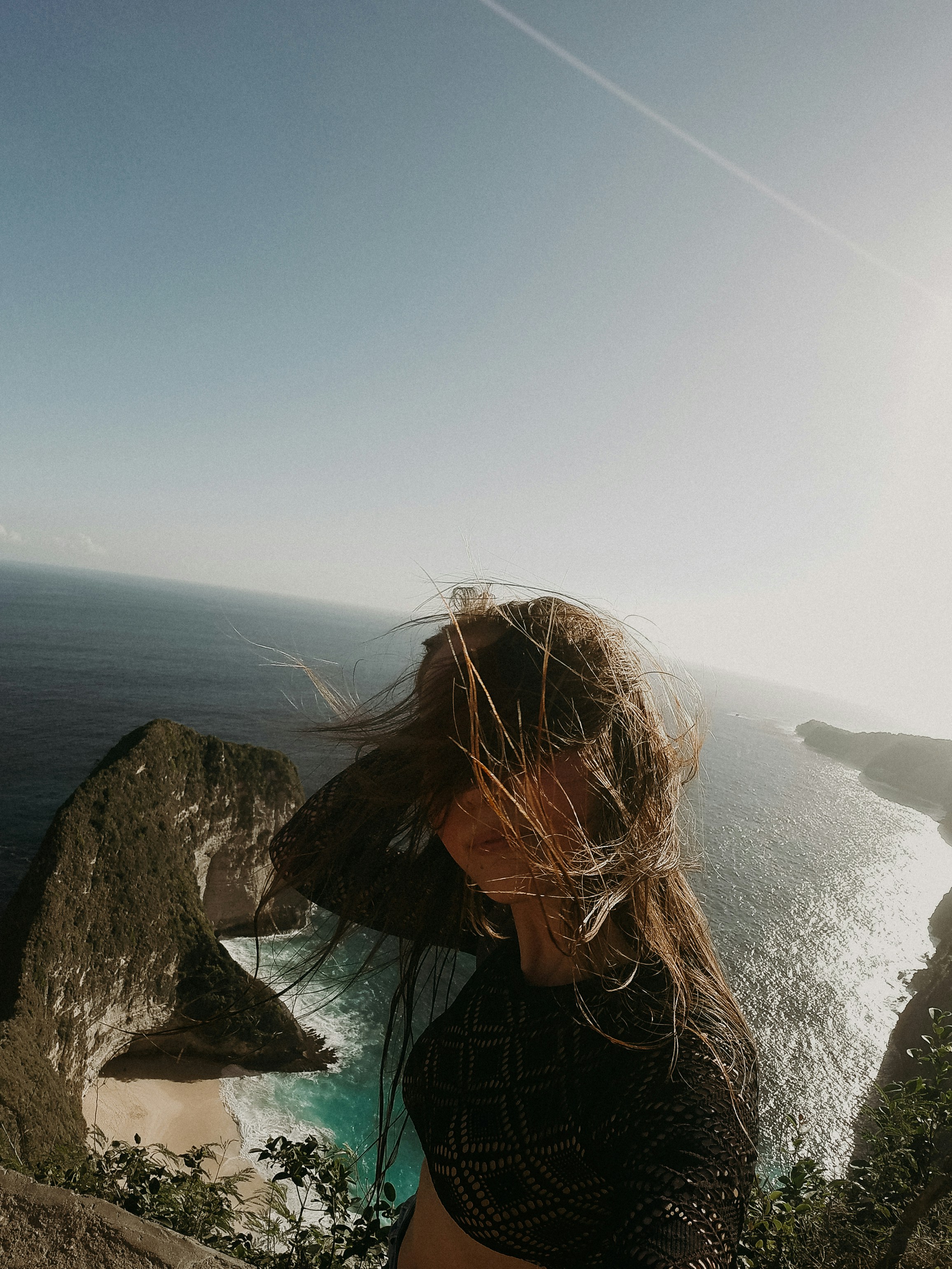 Wind-swept selfie on a cliff overlooking a turquoise ocean, hair blown across the face.