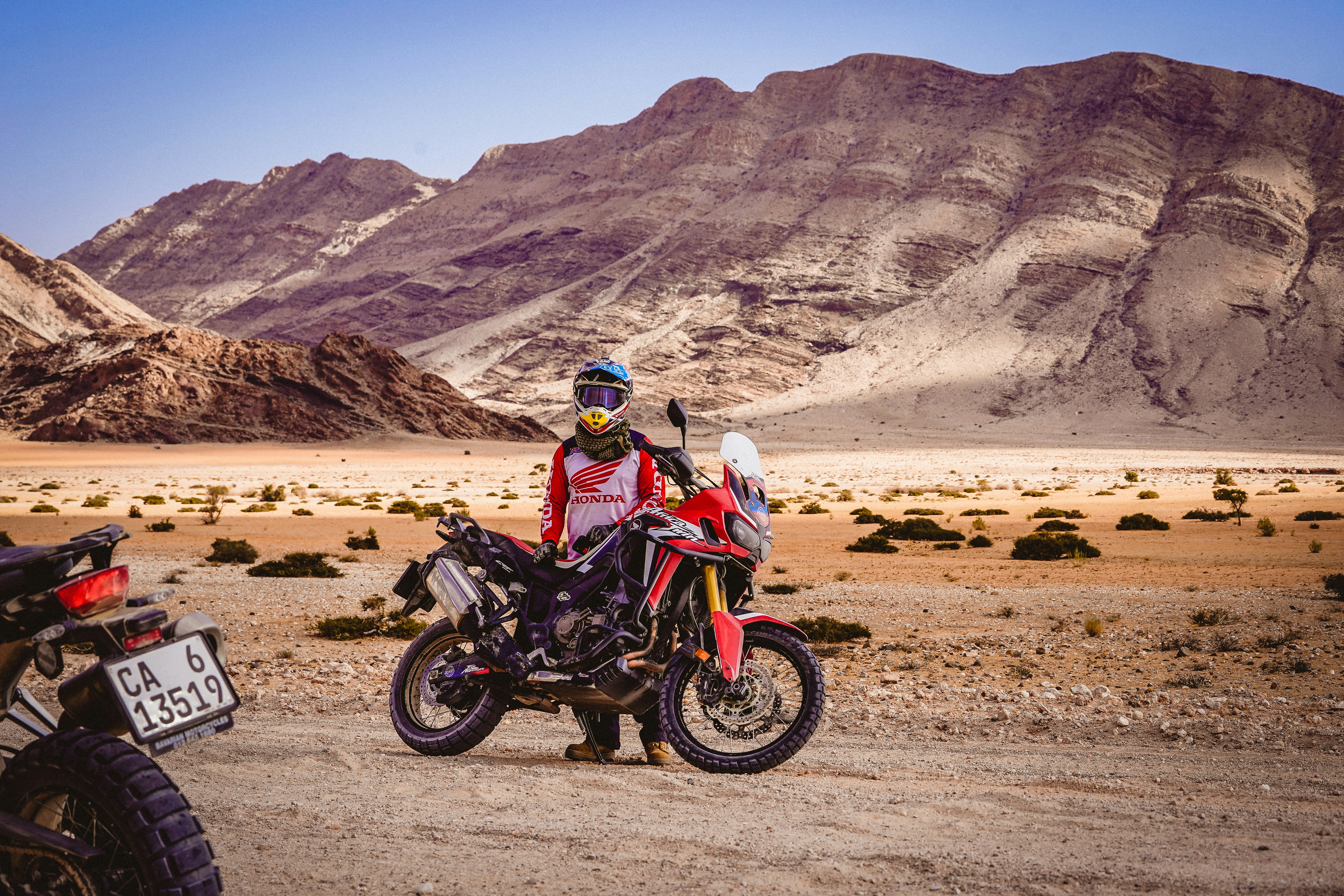 Motorcyclist pauses in a vast desert landscape with rugged hills in the background.