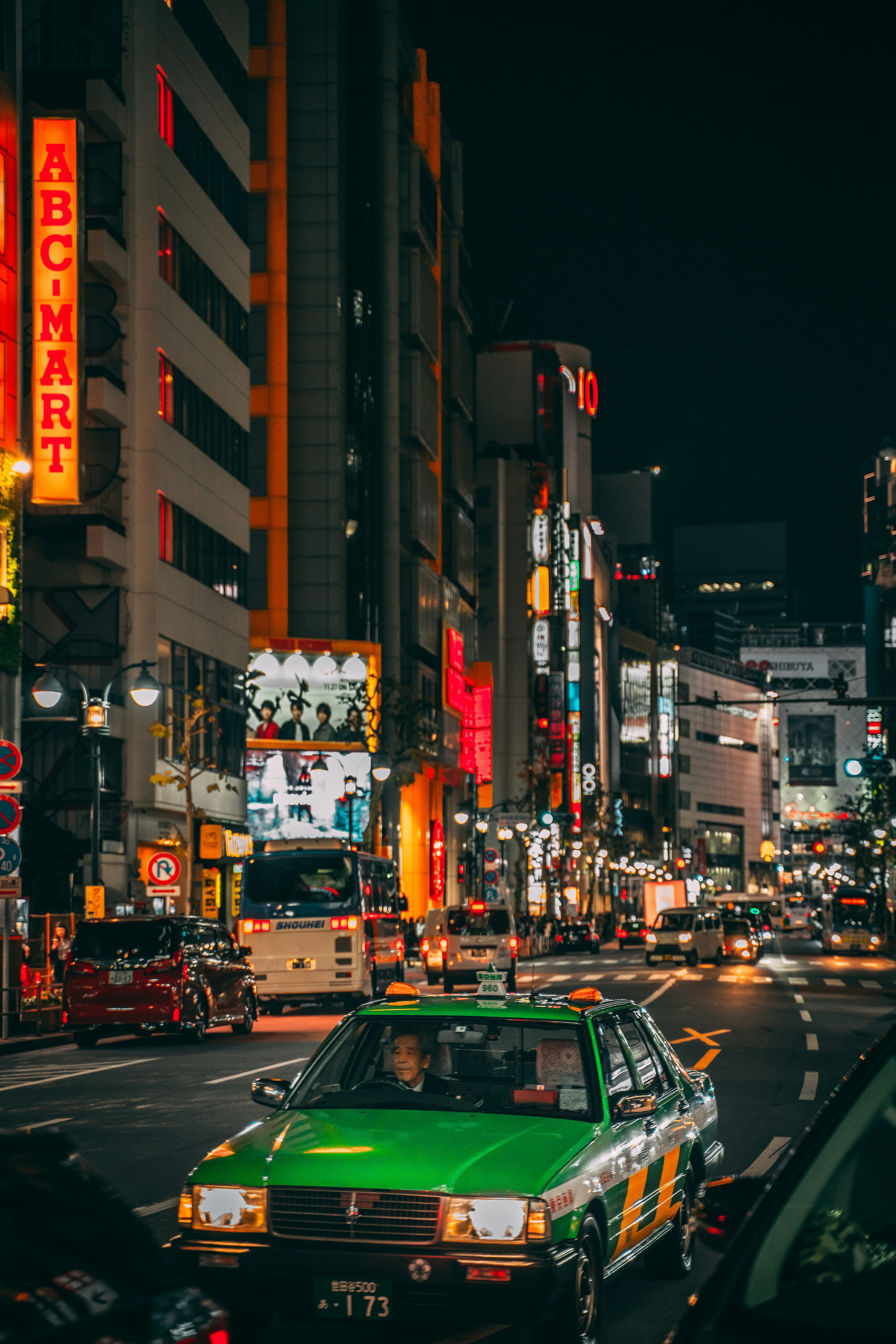 A vibrant city street at night, showcasing a green taxi amidst glowing neon signs and bustling traffic. The scene captures the lively essence of urban nightlife.