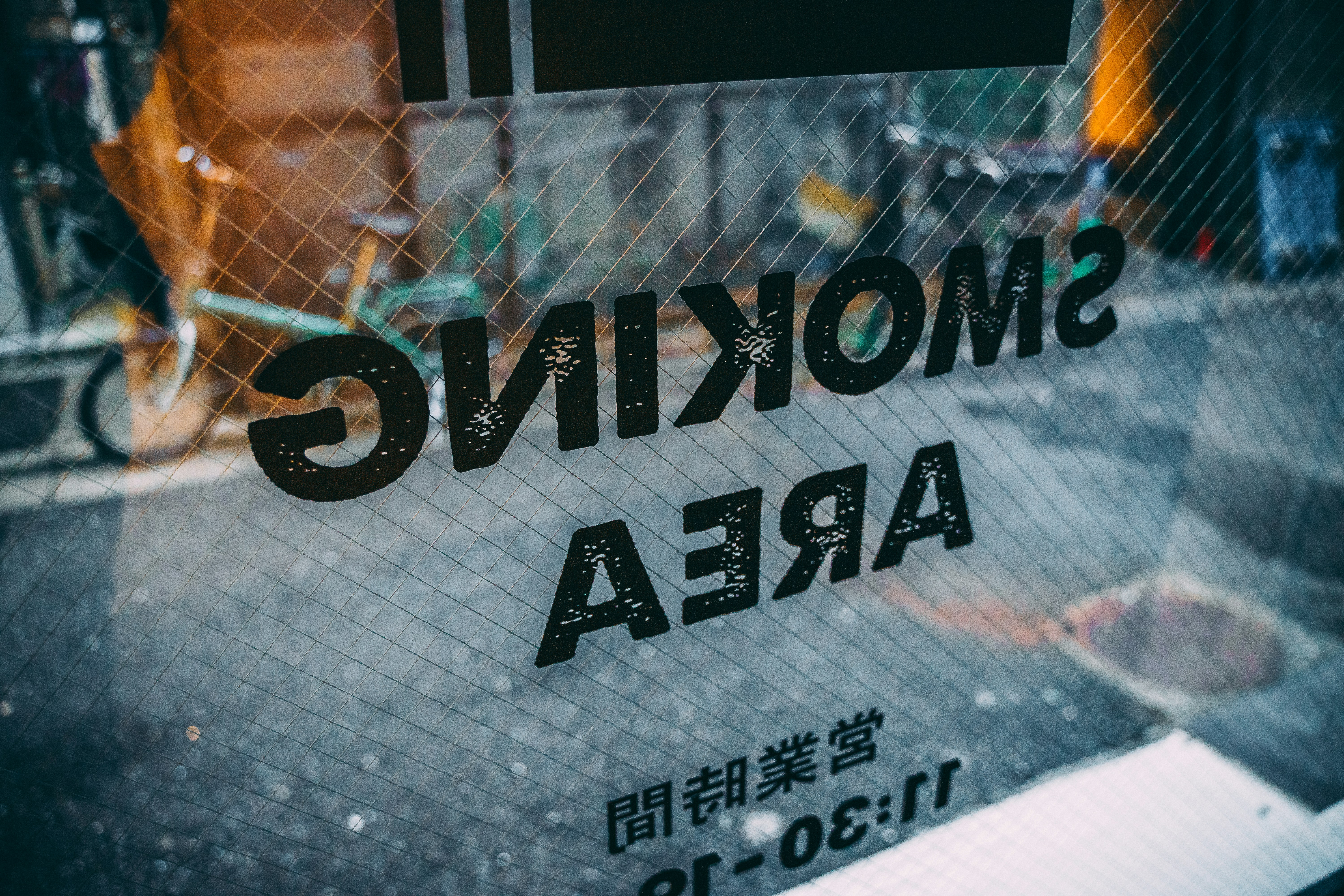Signage indicating a designated smoking area, reflecting urban life through a glass window. The text is partially obscured by the reflection of the surroundings.