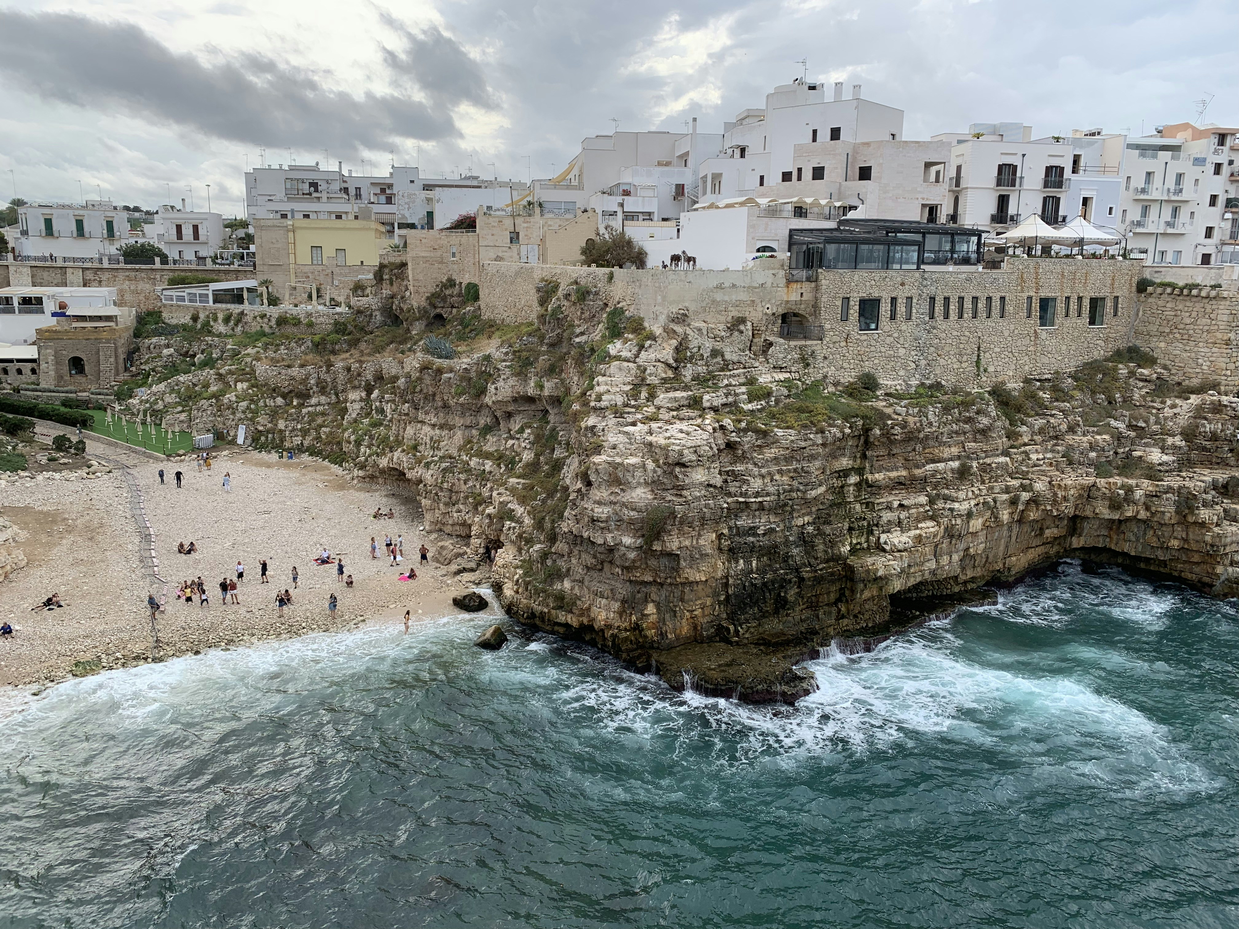 Cliffside town with white buildings overlooking a beach and turquoise waves under a cloudy sky.