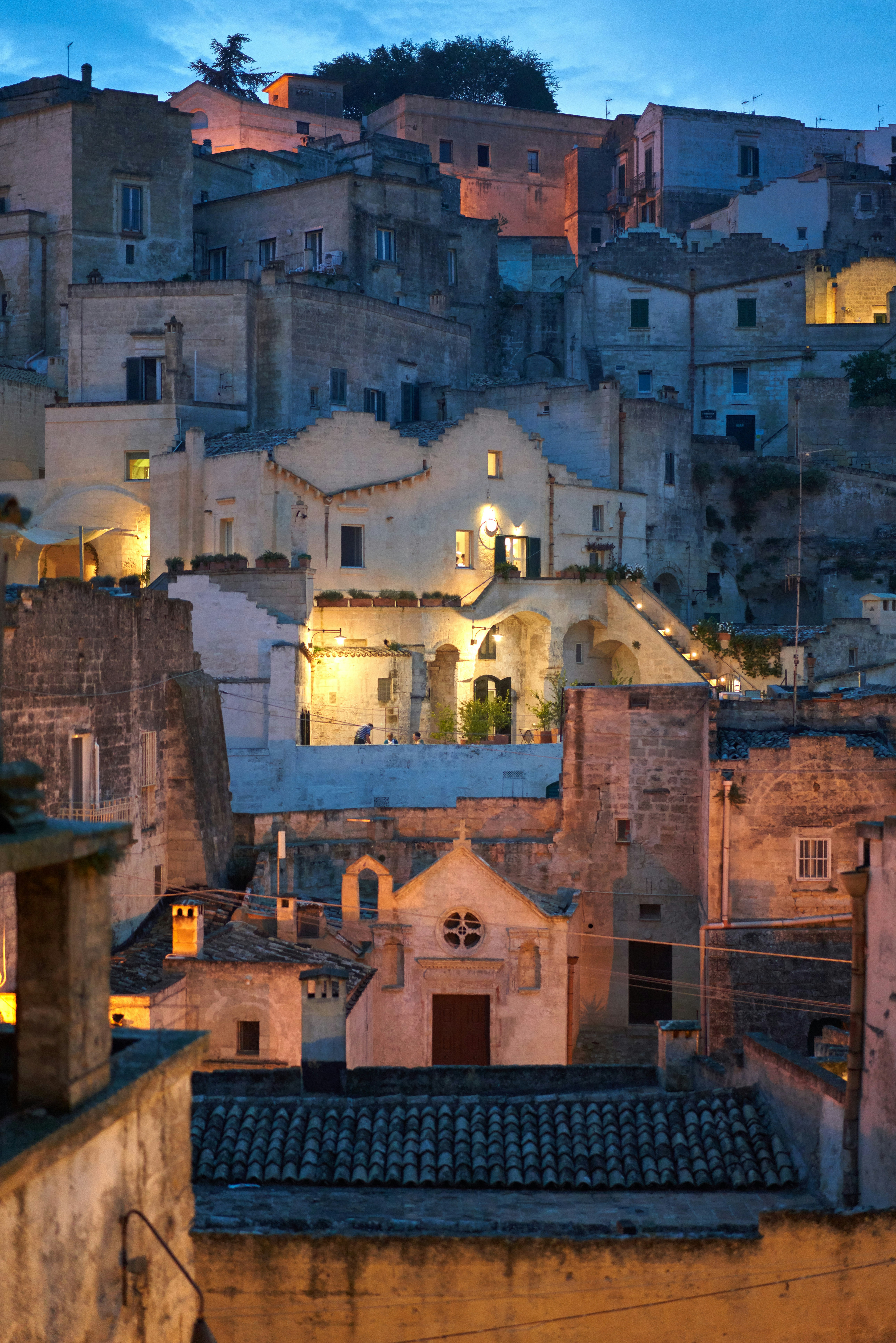 Historic buildings nestled on a hillside, softly illuminated against a twilight sky. The scene captures the charm of ancient architecture and the interplay of light and shadow.