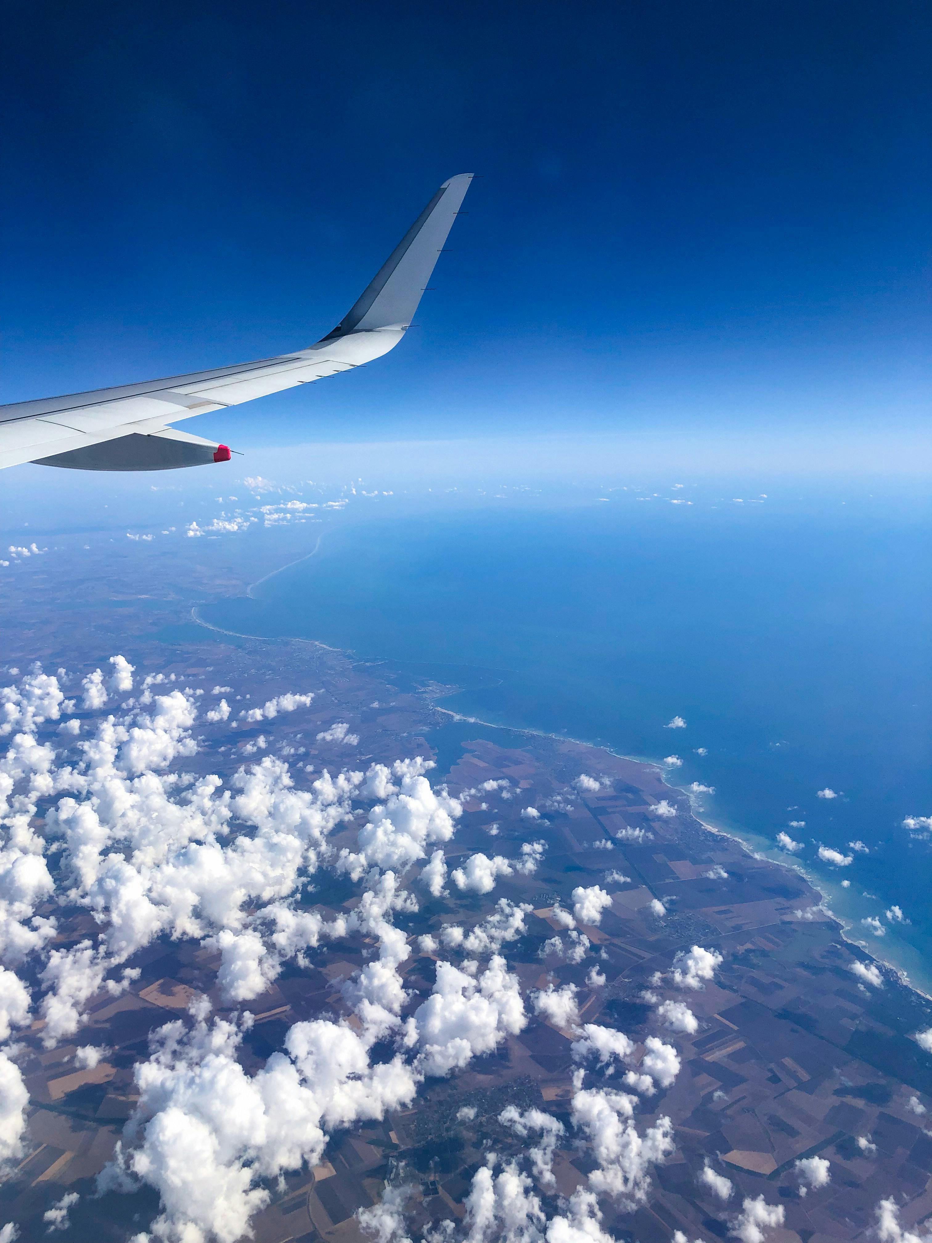 Airplane wing extending above a patchwork of fields and clouds, with a vast ocean in the distance.