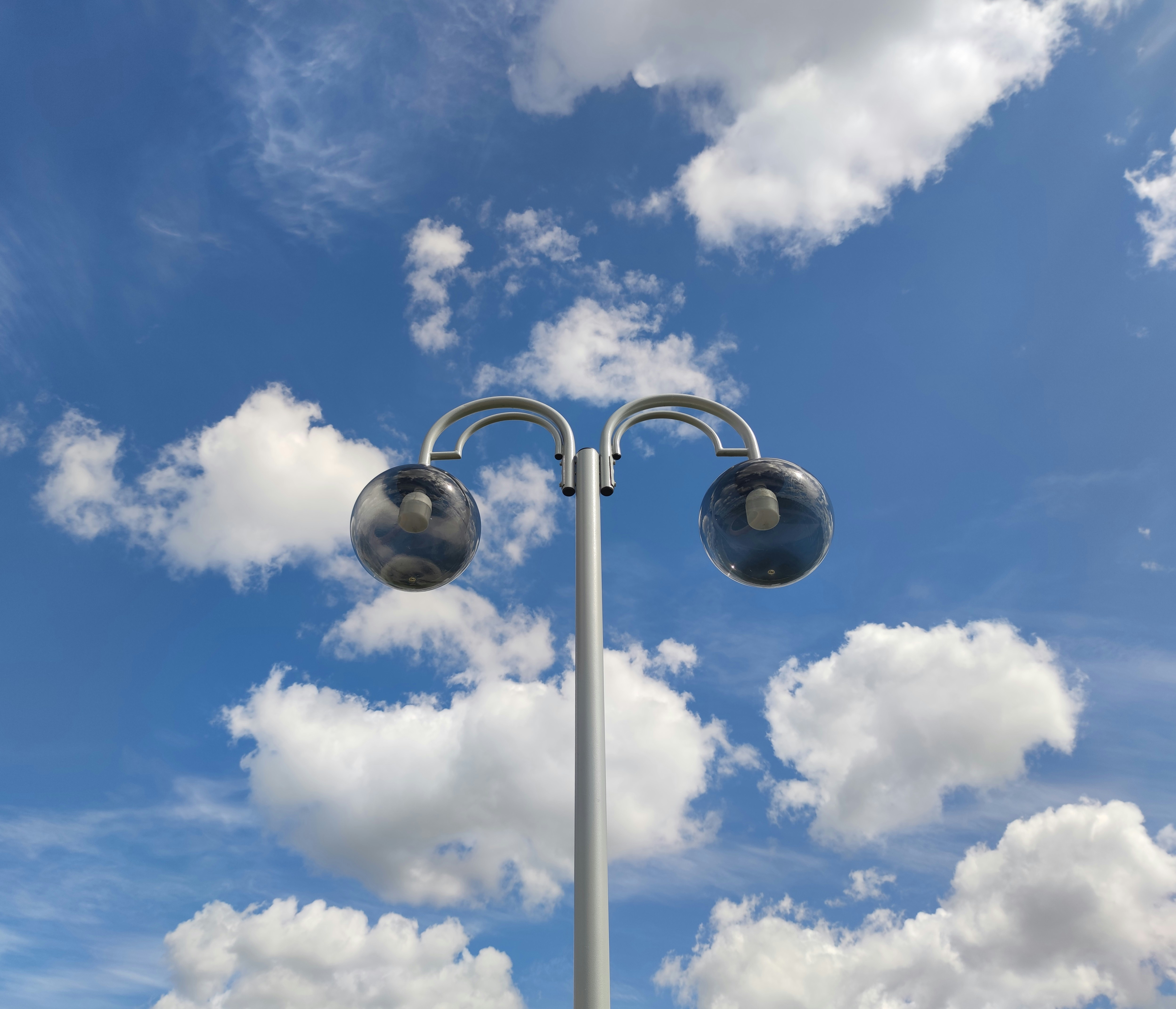 Two glass globes of a street lamp rise against a bright blue sky dotted with clouds. The composition emphasizes vertical geometry and the crisp contrast between metal, glass, and sky.