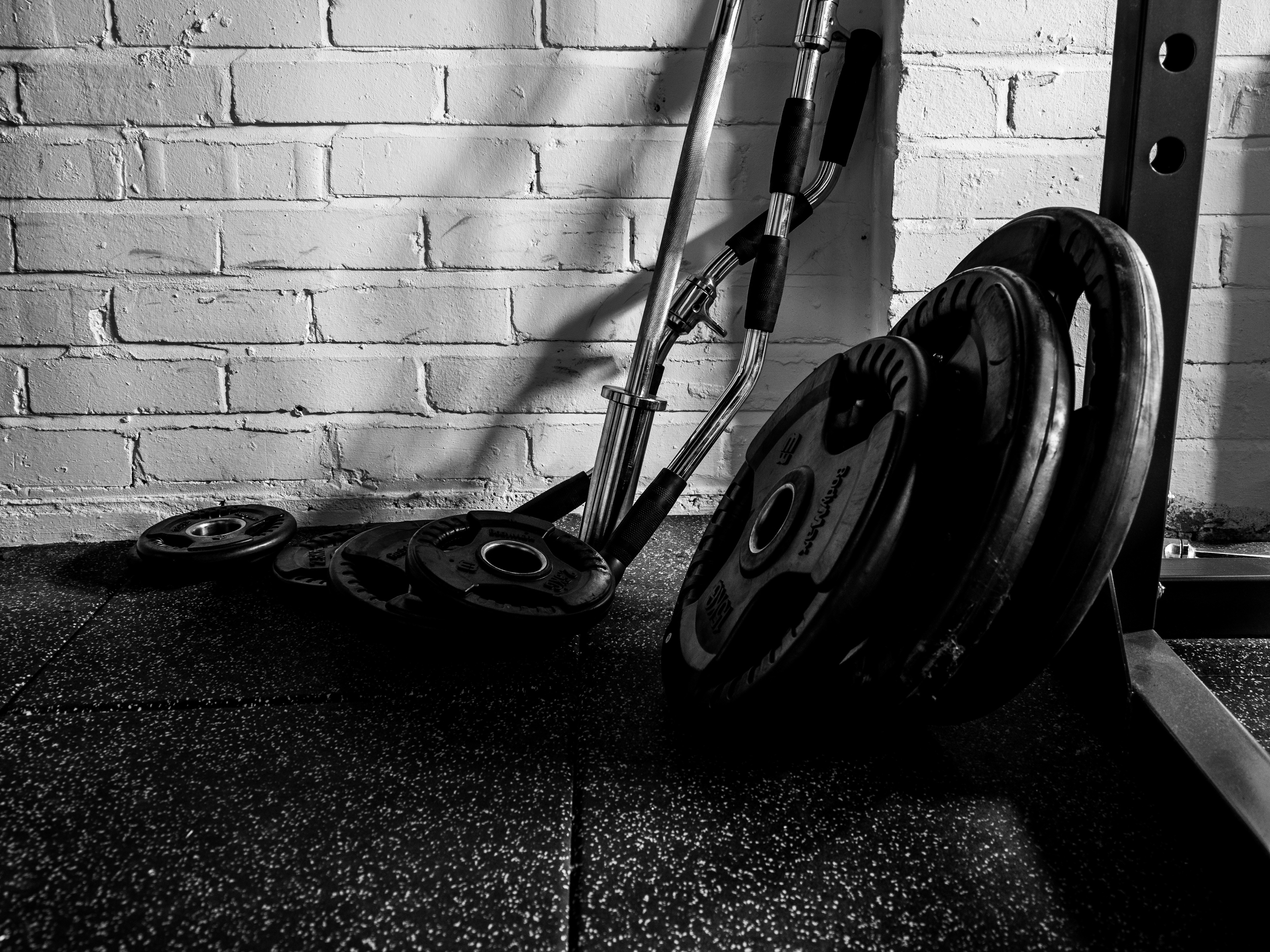 A collection of weight plates and a barbell resting against a textured black floor and white brick wall, emphasizing the essence of strength training.