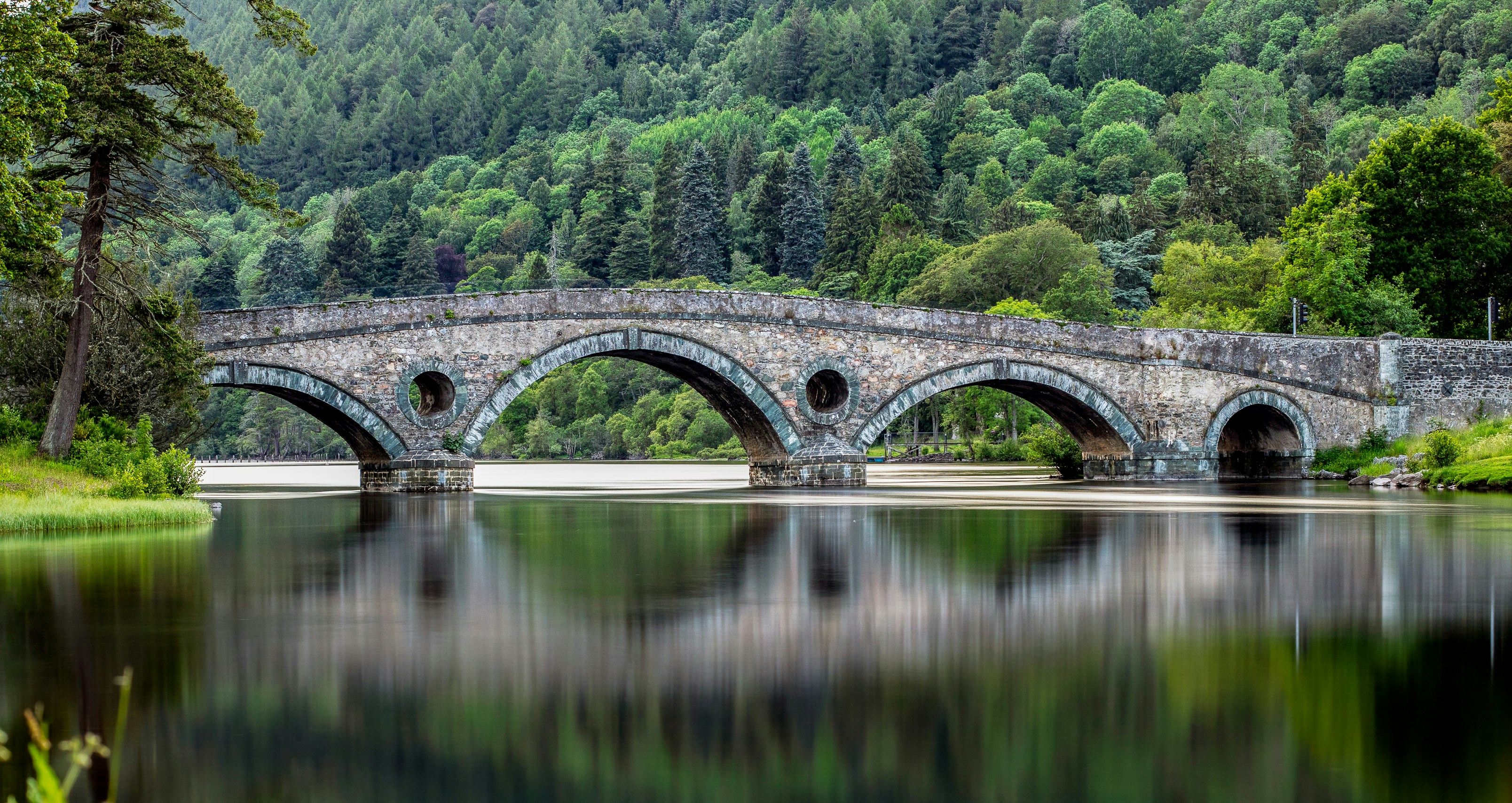 The old bridge, Kenmore, Perthshire, late in the evening with a slow shutter.  The sun had already dropped below the horizon, with the green, tree-lined hills in shadow. | gray concrete bridge over river
