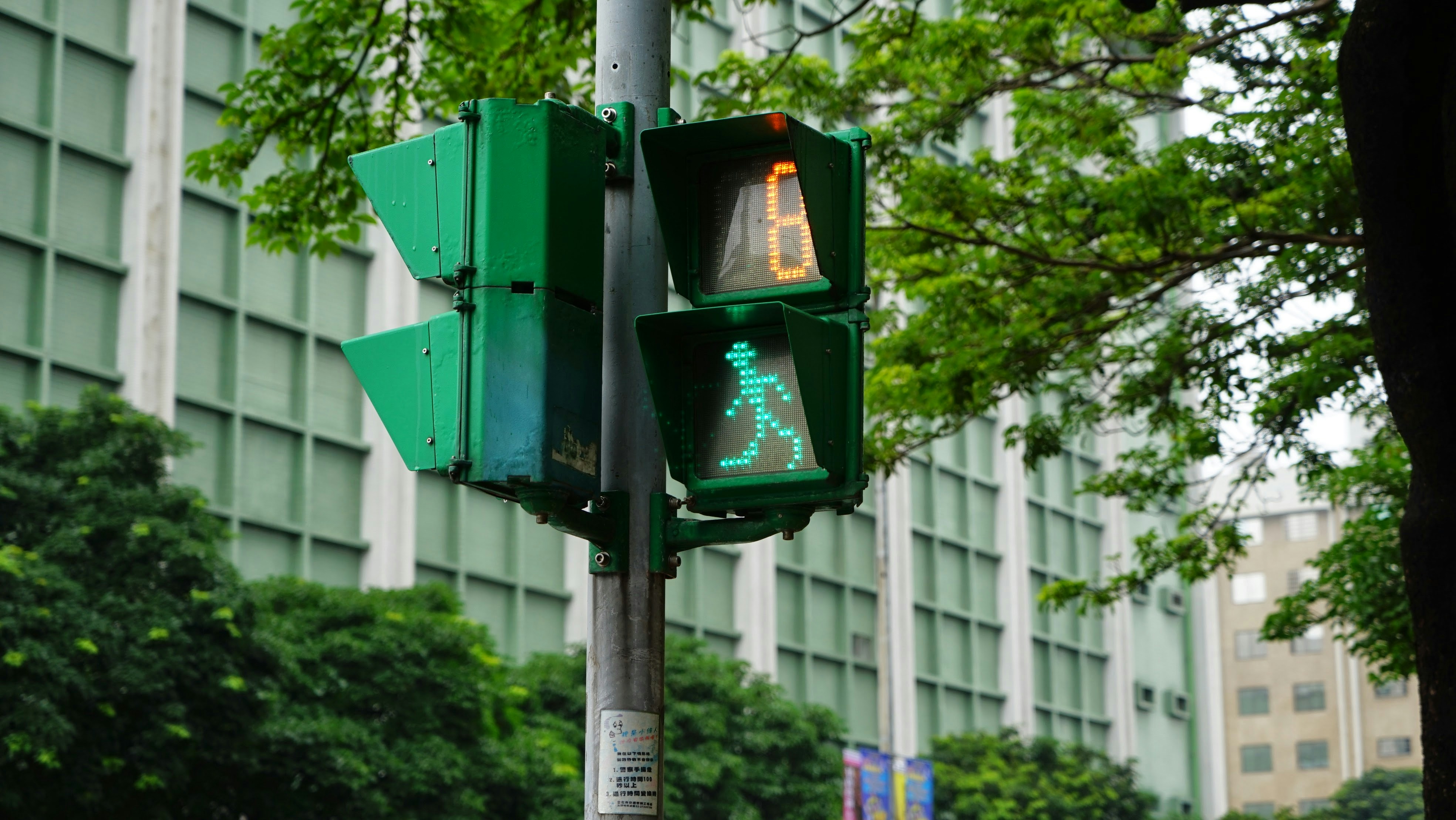 Traffic signal displaying a green pedestrian symbol against a backdrop of urban architecture and foliage.