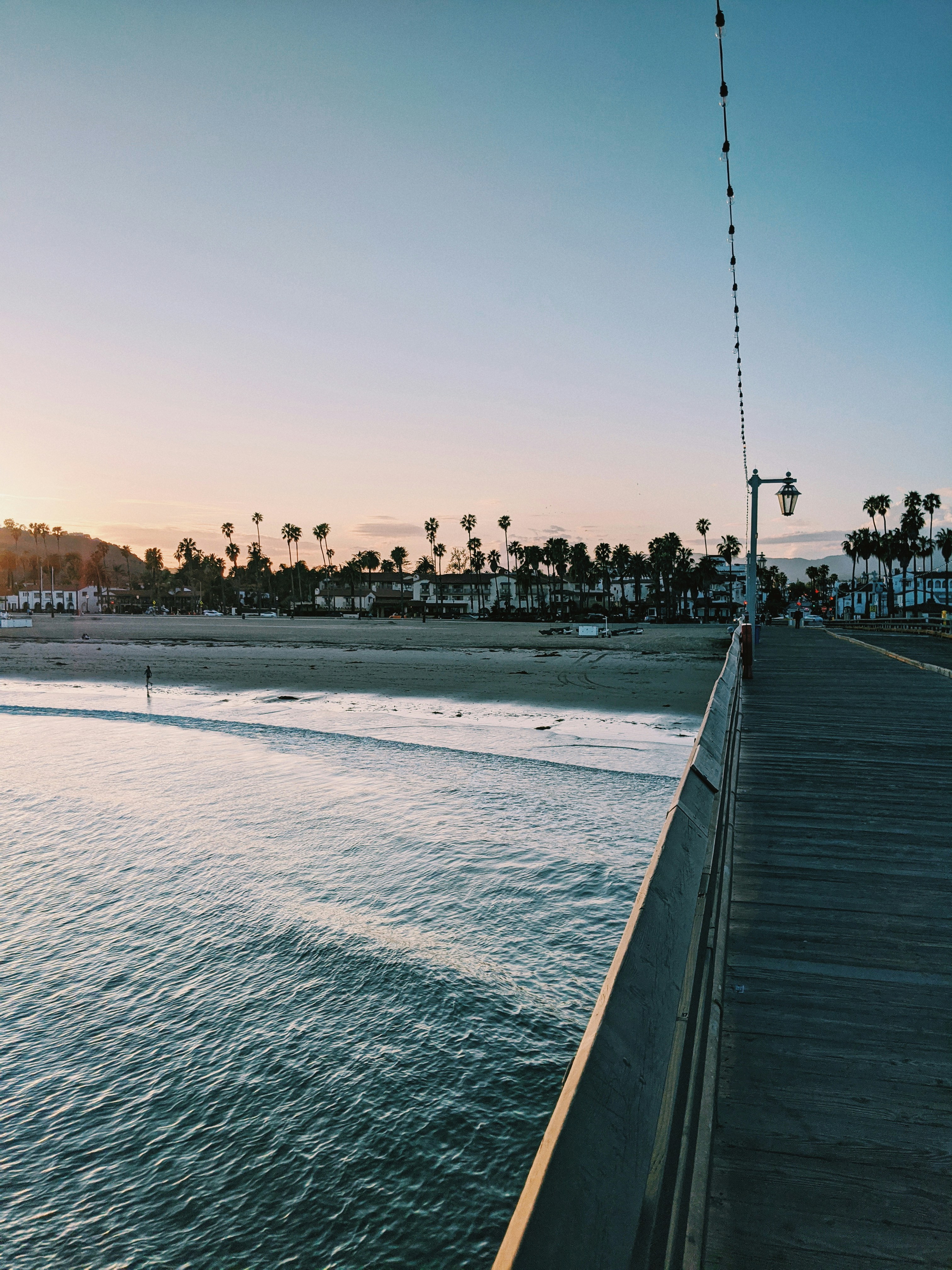 Wooden pier leading towards a tranquil beach at sunset, with palm trees lining the shore and soft waves lapping at the sand.
