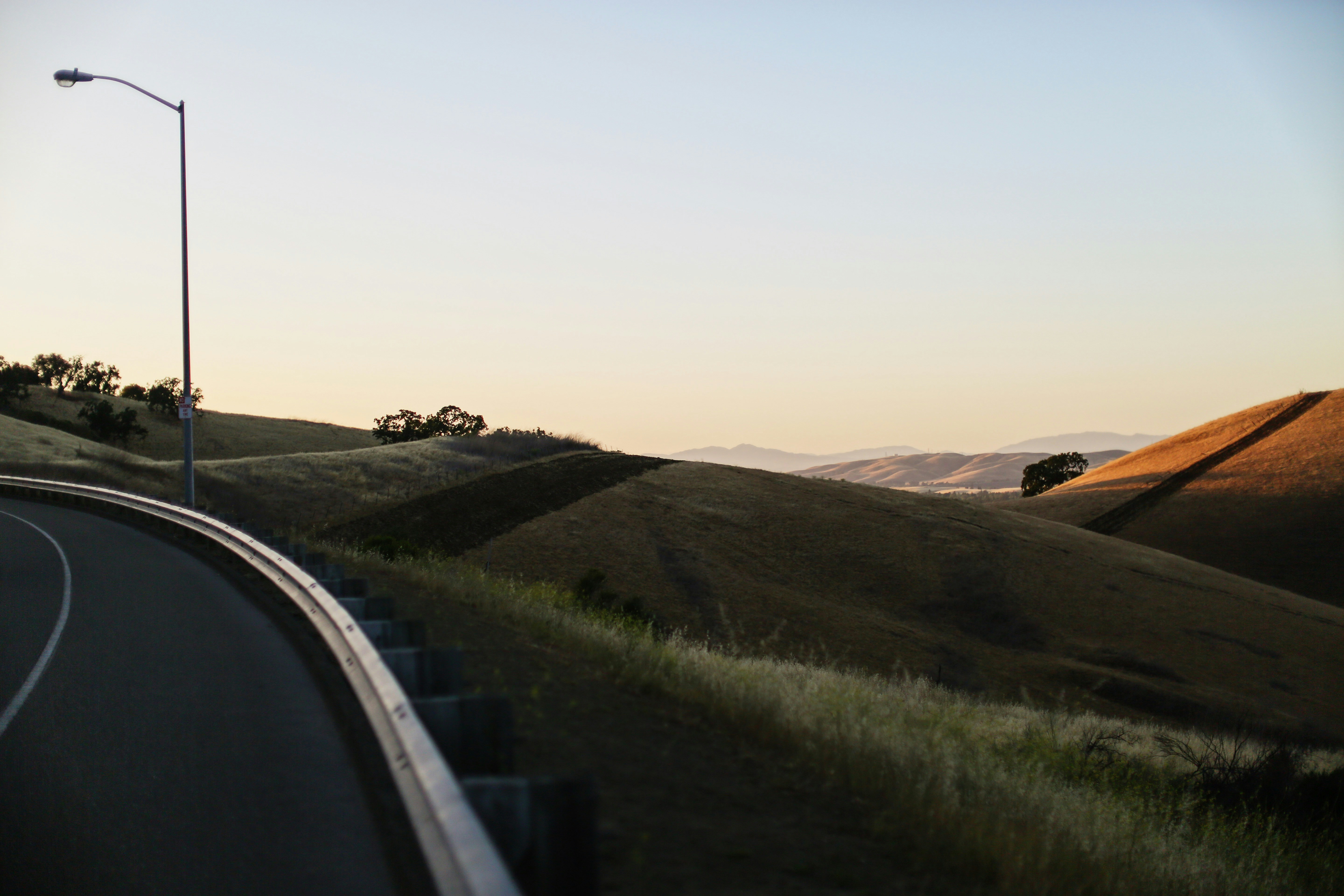 Black car traveling along a winding road beside sunlit hills and green fields at dusk.