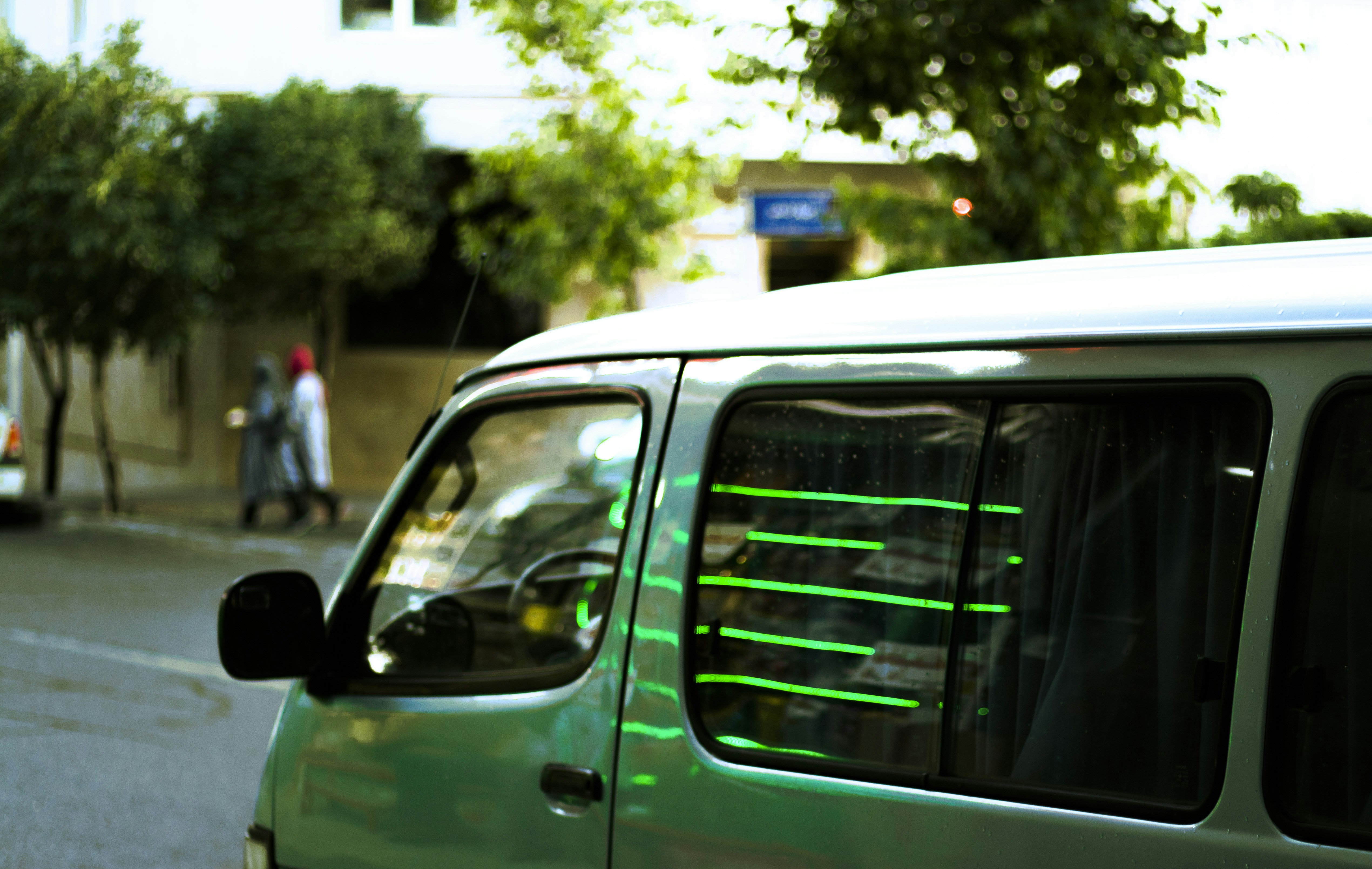 A green van parked on a city street, reflecting neon lines against its window, with pedestrians in the background. The scene captures a moment of urban life.