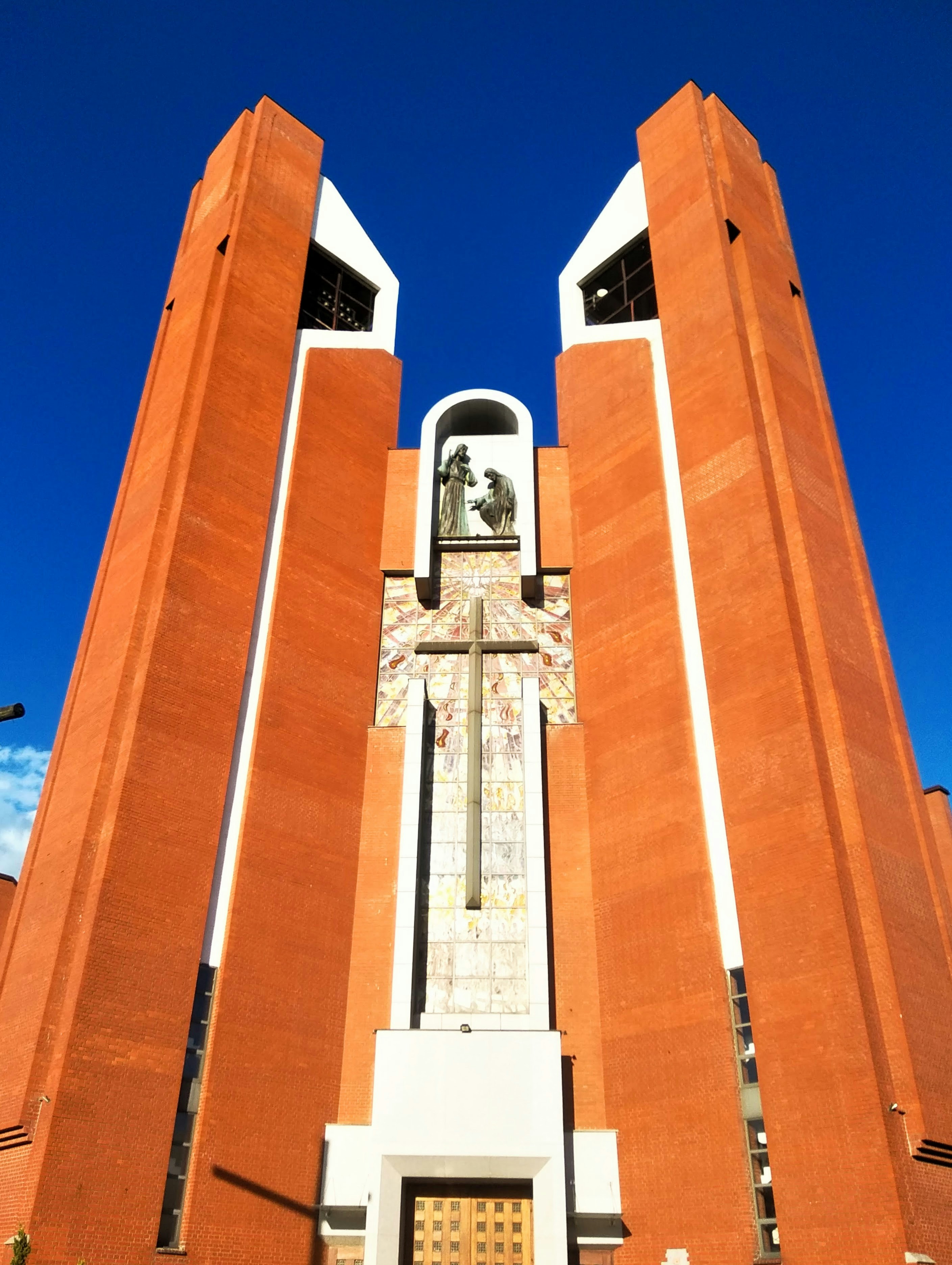 Striking red brick church with a prominent cross and statue, set against a clear blue sky. The design showcases a blend of modern and traditional elements.