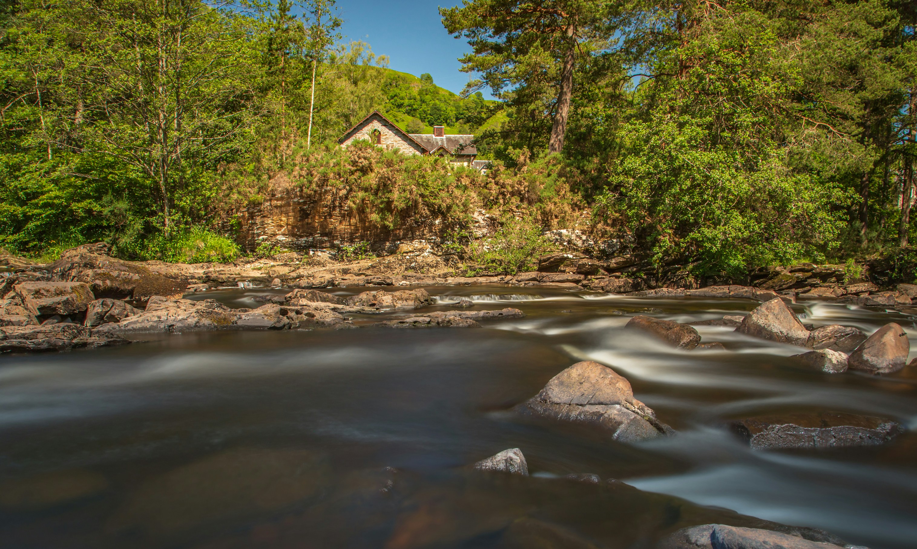 A tranquil riverside scene featuring a cozy cottage nestled among lush greenery, with smooth water flowing over rocks in the foreground.