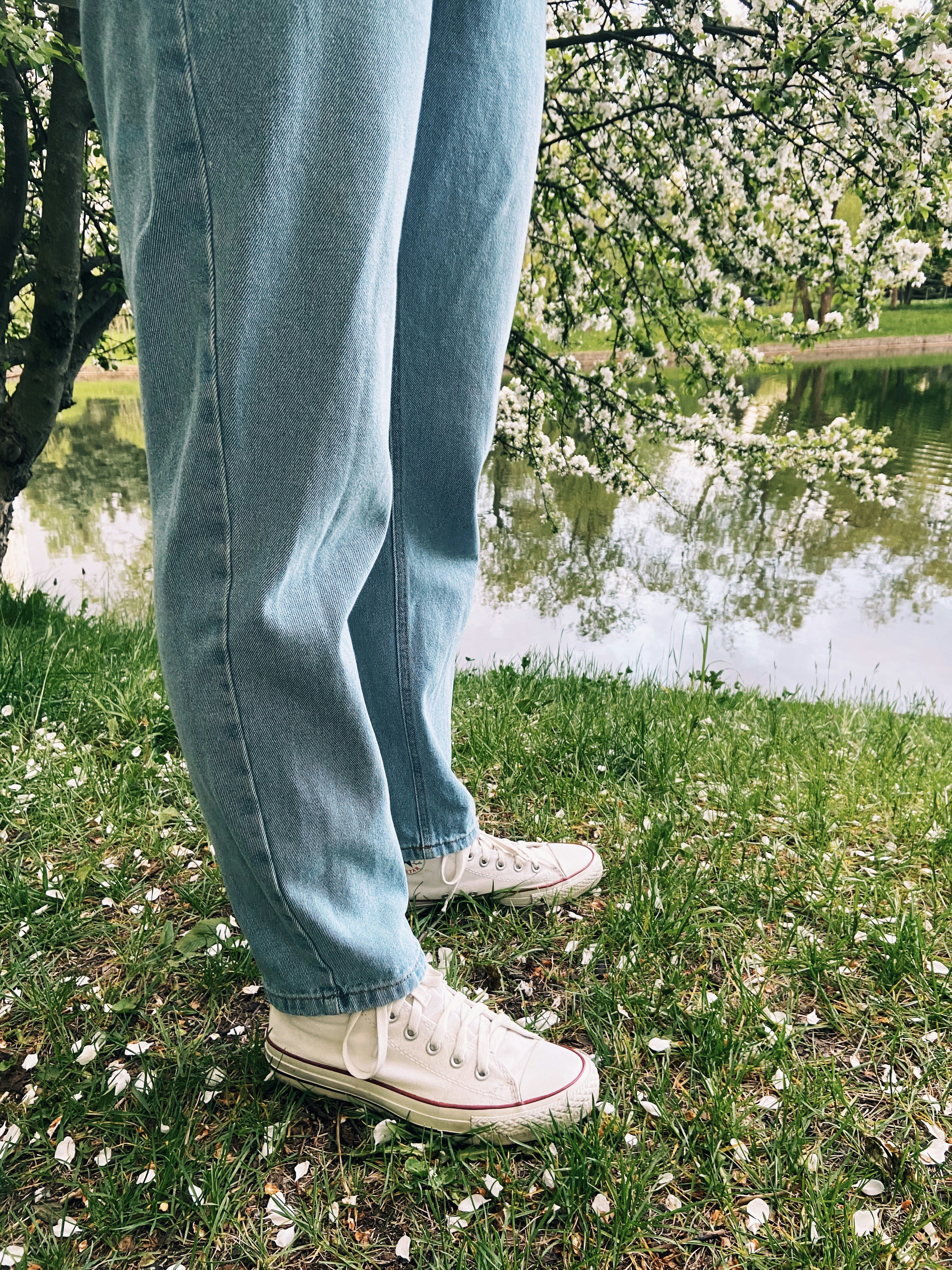 Light blue jeans and white sneakers stand amidst blooming flowers by a tranquil pond.