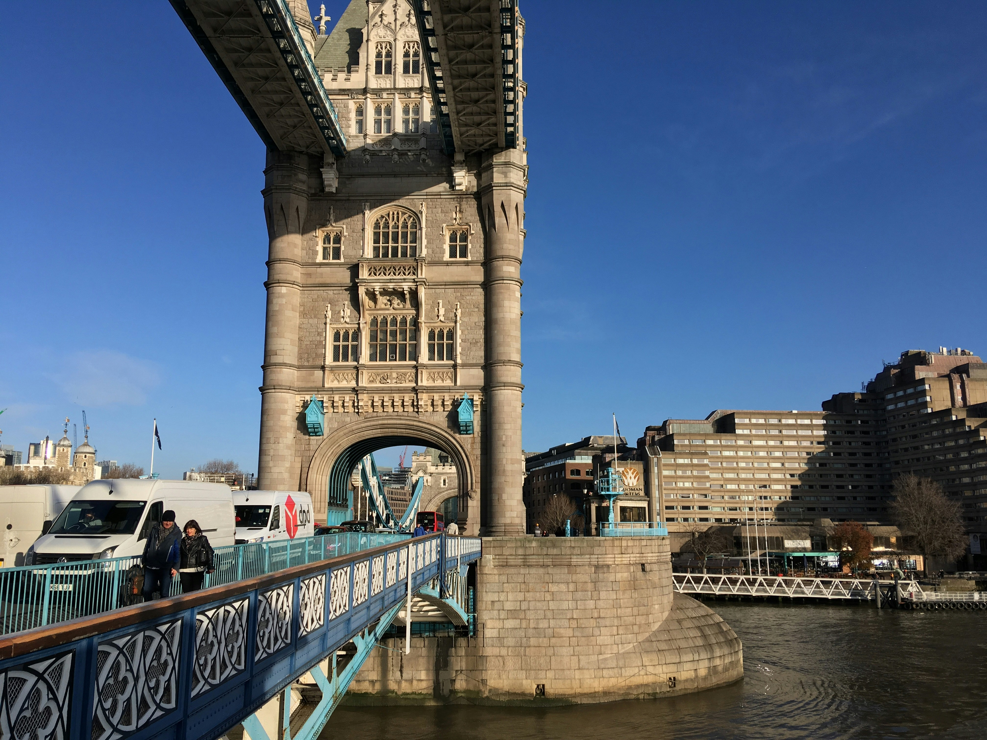 Tower Bridge rises over the Thames with pedestrians along the blue walkway under a bright blue sky.