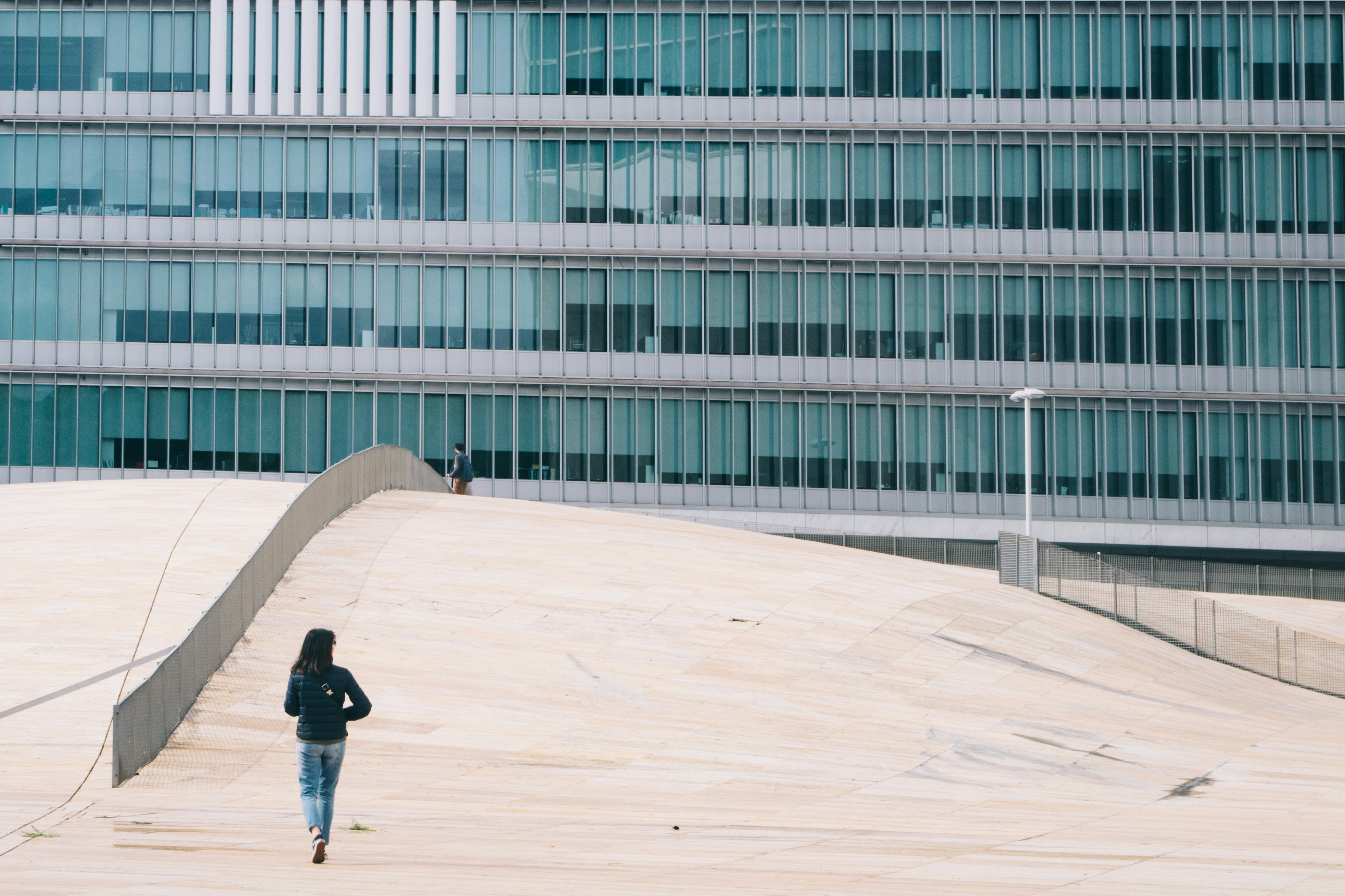 A solitary figure walks along a gently curved pathway leading to a modern building, showcasing the interplay between architecture and nature.