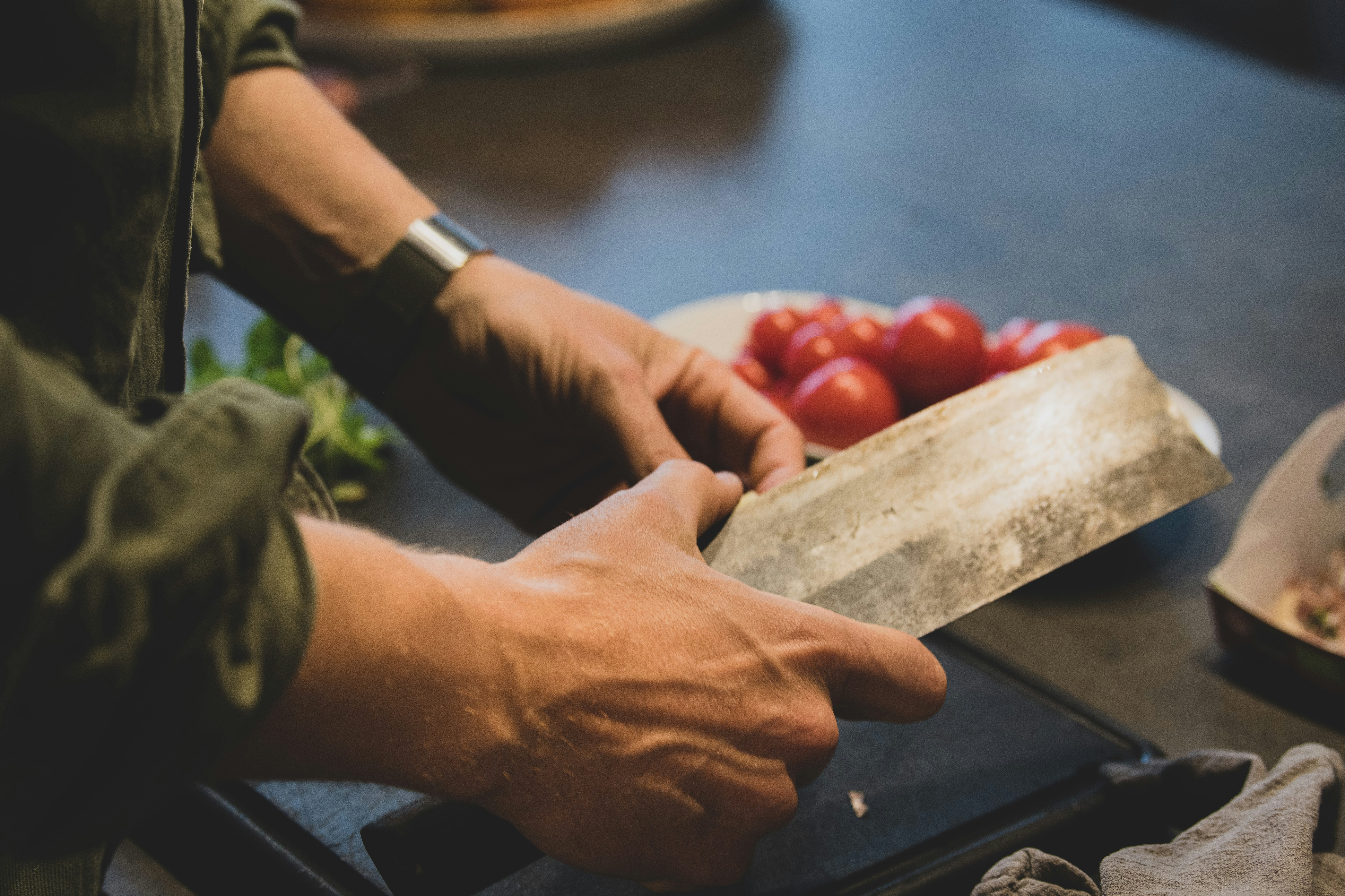 Hands skillfully wielding a cleaver over a cutting board with ripe tomatoes in the background.
