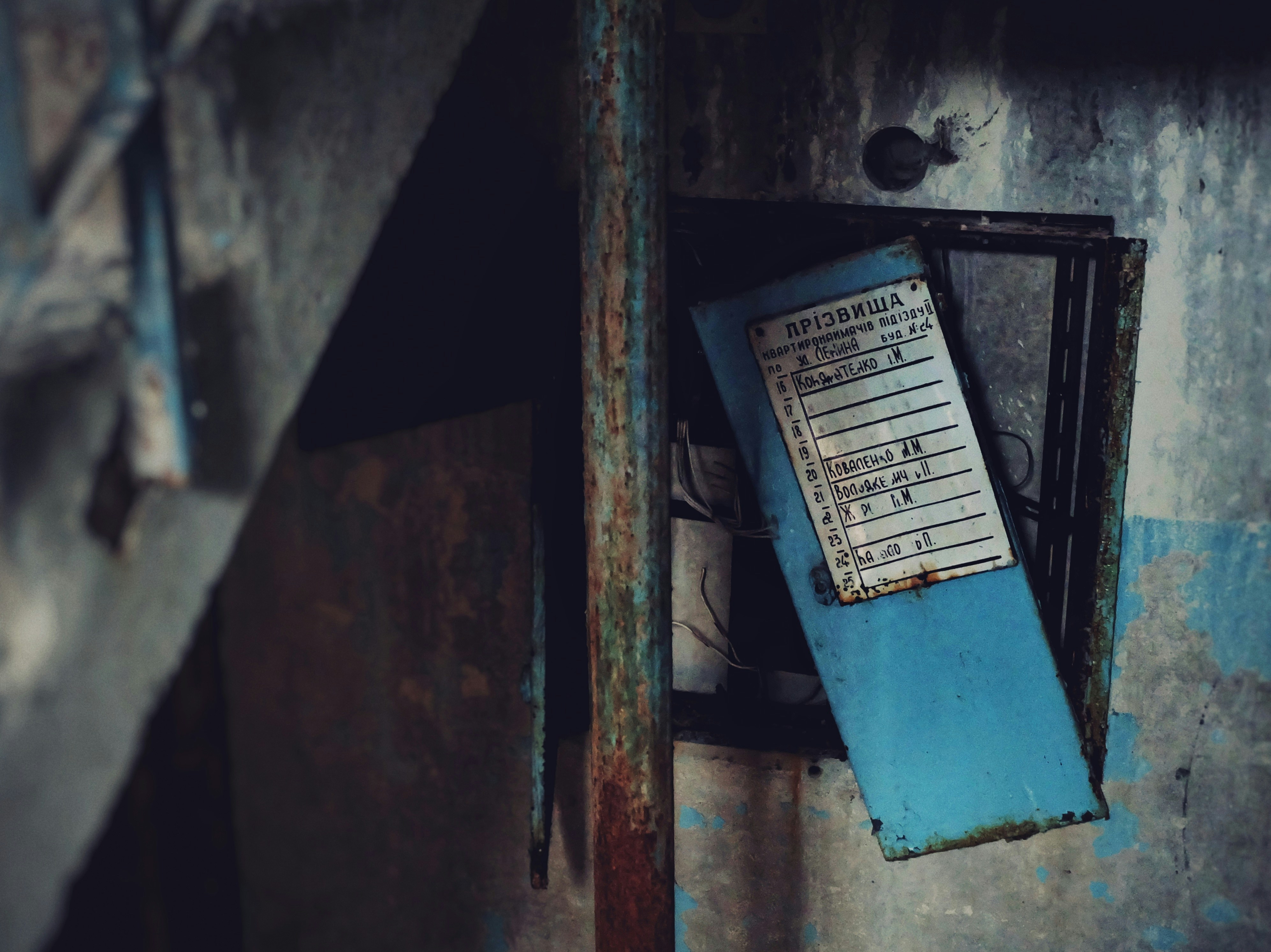 Rusty blue utility box hanging askew in a dimly lit stairwell, revealing remnants of past usage. The peeling paint and shadows evoke a sense of nostalgia.