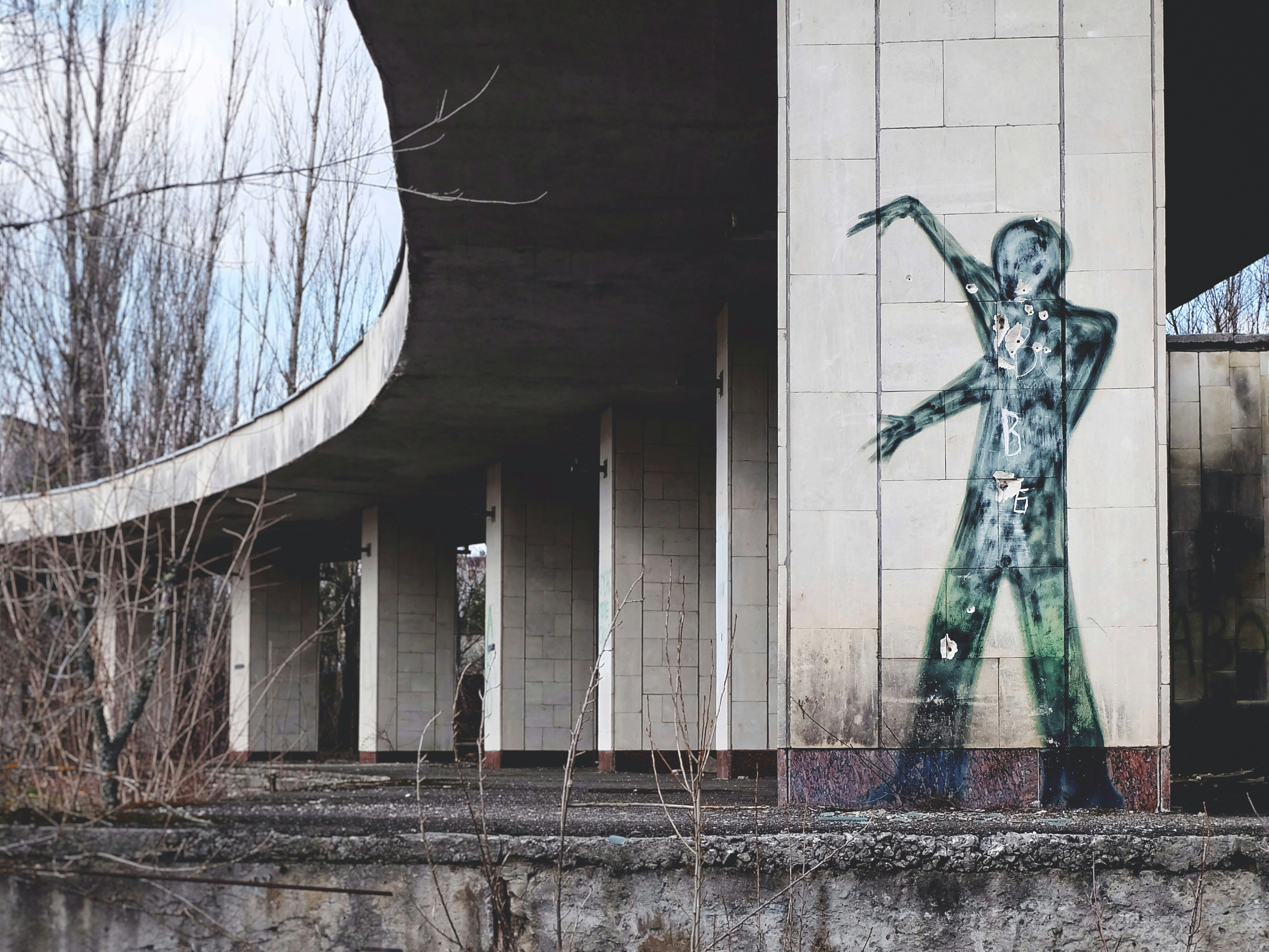 A ghostly figure painted on a crumbling wall amidst an abandoned architectural structure, surrounded by overgrown vegetation.