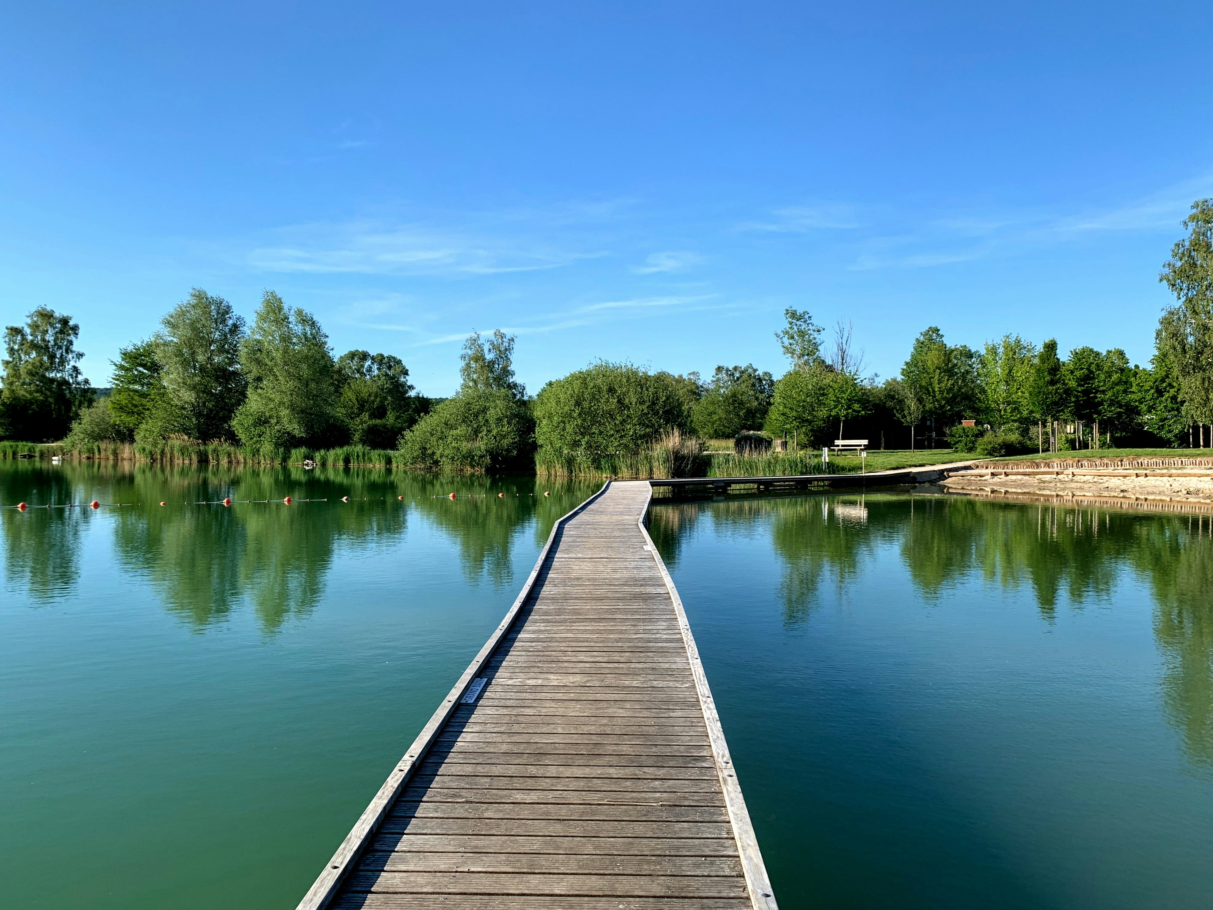 Wooden walkway extending into a tranquil lake surrounded by lush greenery and reflections of trees in the water.