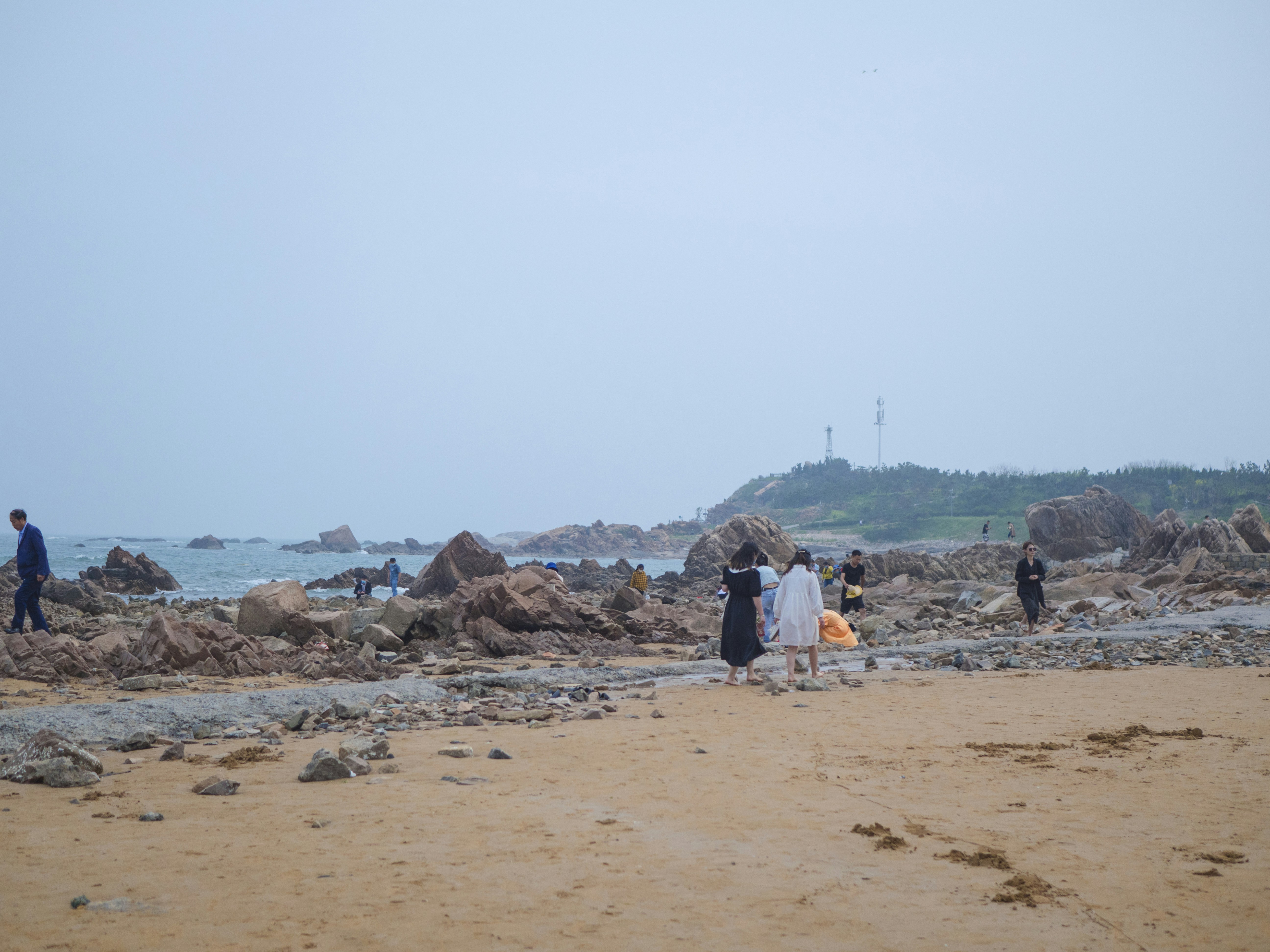 Two figures in white traverse a windswept rocky beach under a pale sky
