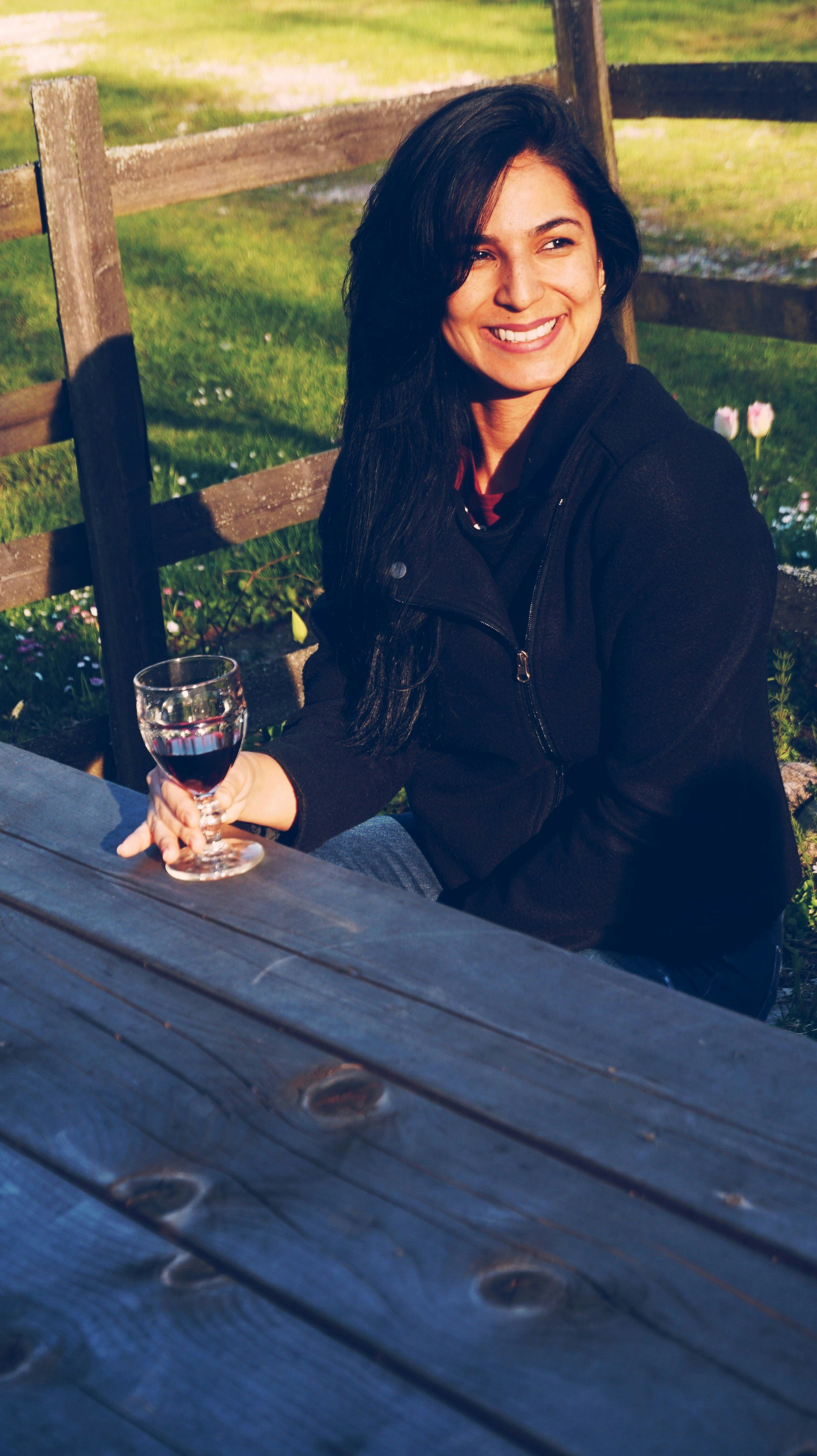 Woman enjoying a glass of purple drink while seated at a rustic wooden table in a serene outdoor setting.