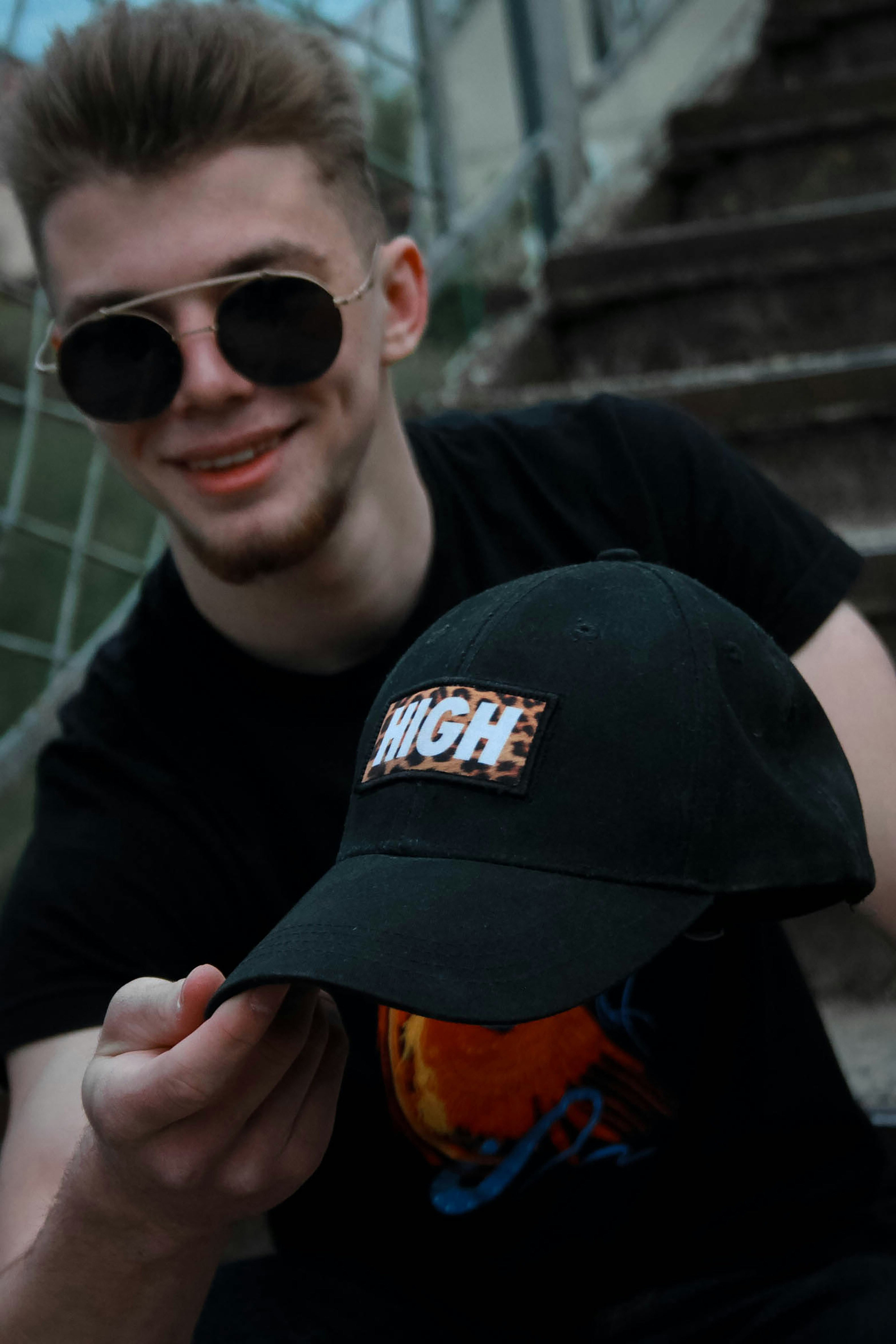 A young man in sunglasses playfully presents a black cap with a distinctive patch while sitting on stone steps.