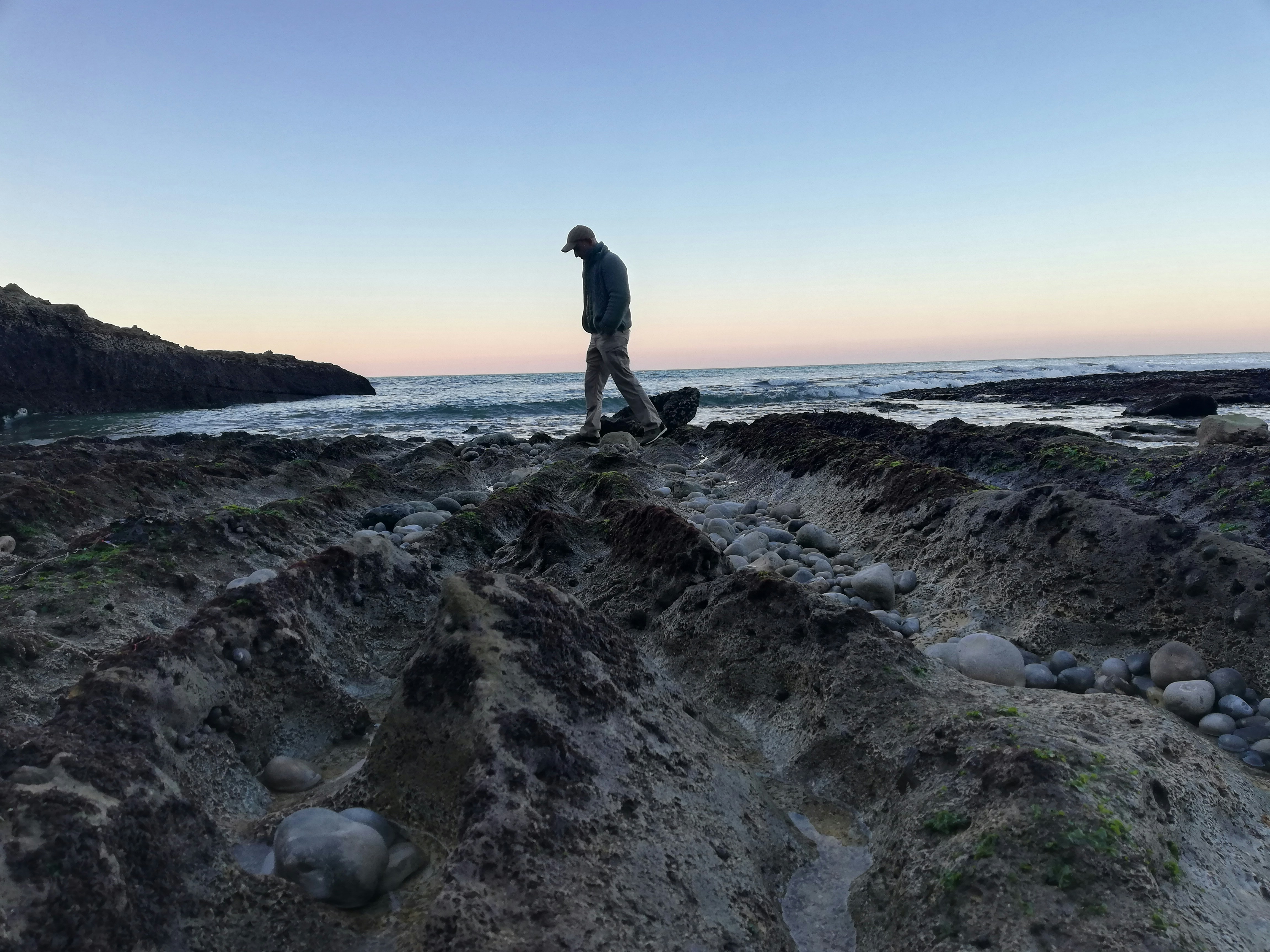 Person walking along rocky shoreline at dusk, with gentle waves lapping at the shore. The soft gradient of the sky transitions from blue to warm hues.