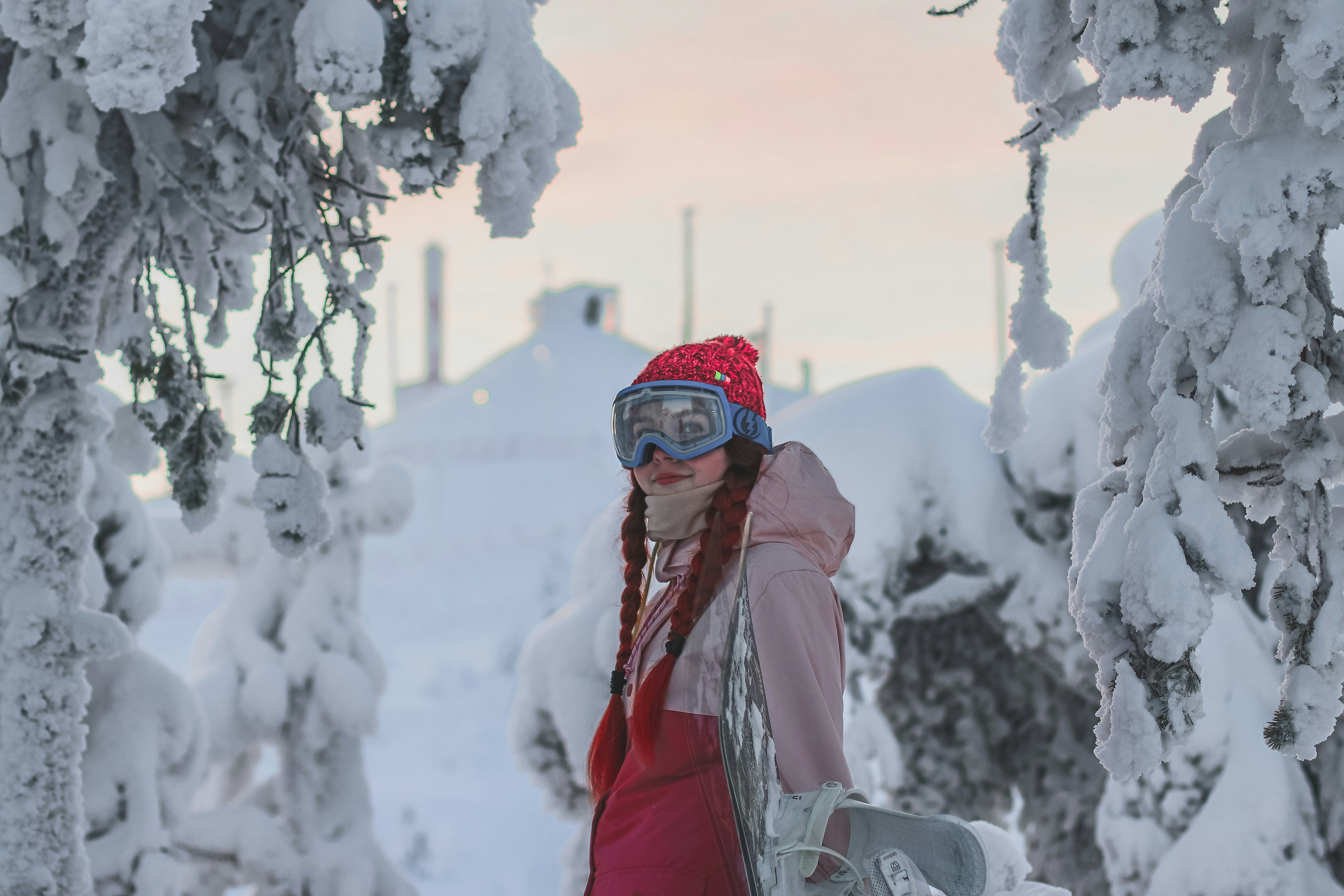 Snowboarder adorned in vibrant gear stands amidst a snowy landscape, framed by frosted trees. The soft light of dawn casts a serene glow over the scene.