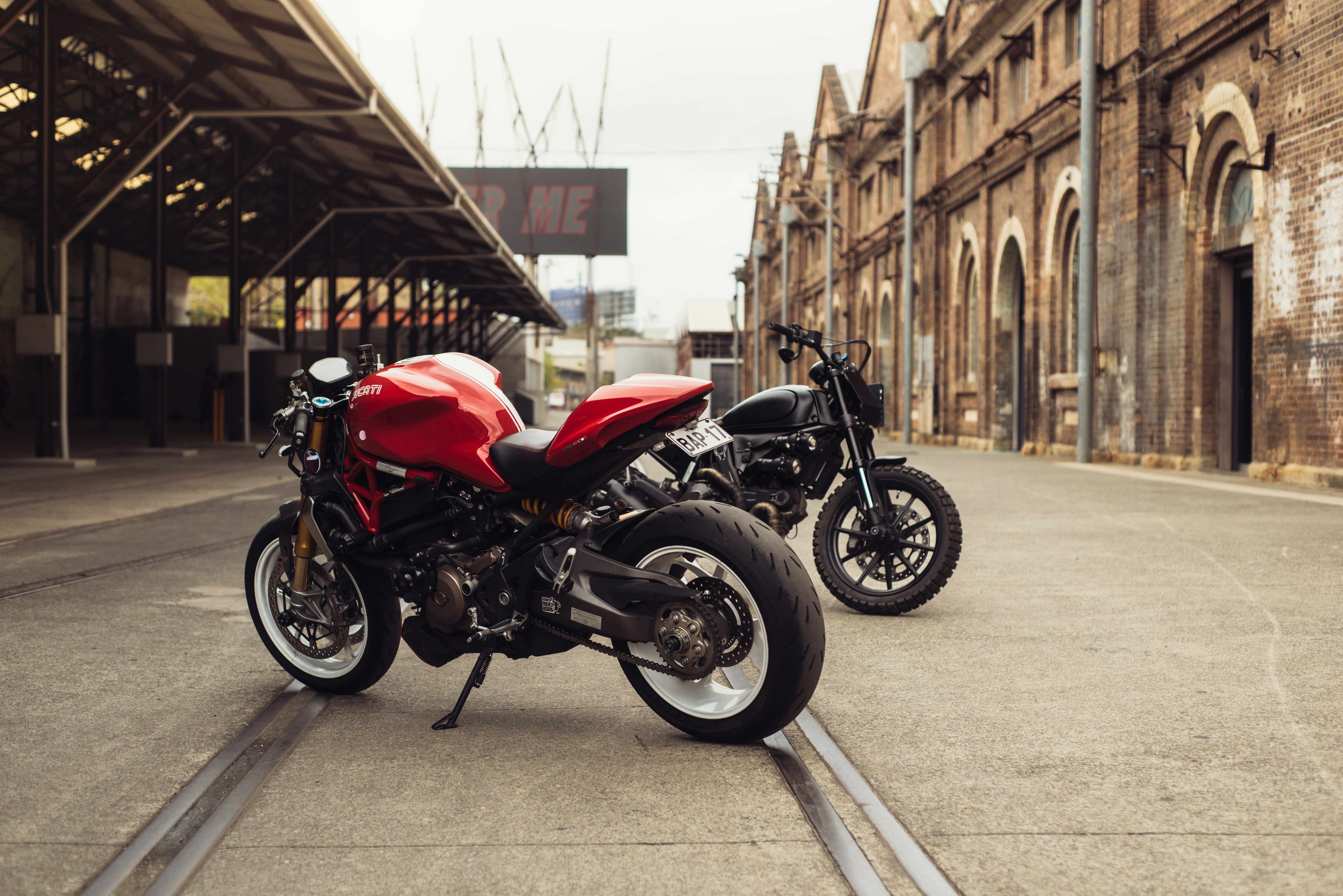Two motorcycles, one red and one black, positioned on railway tracks in an industrial setting, showcasing their sleek designs against a backdrop of heritage architecture.