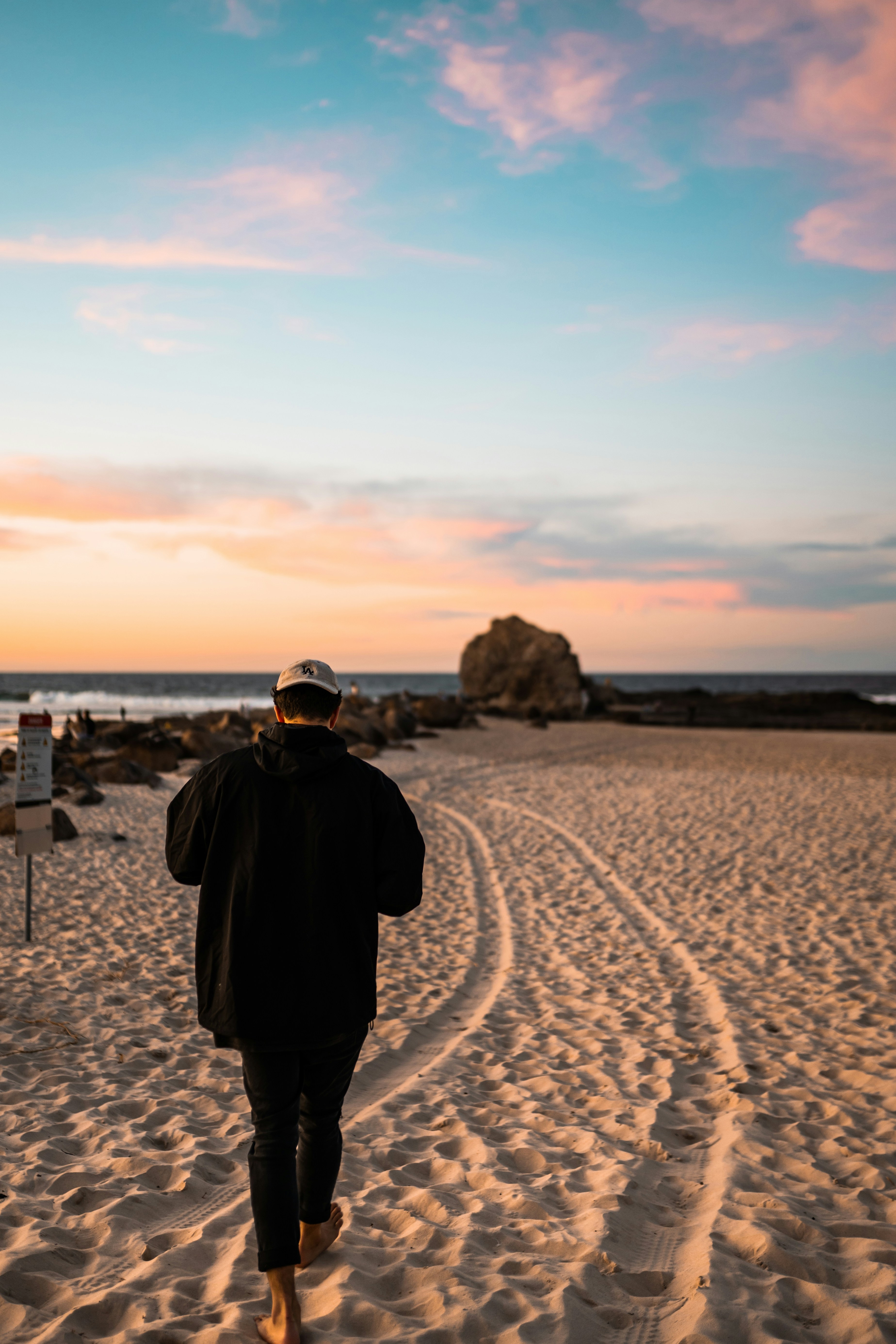 A figure walks along a sandy beach, leaving footprints in the soft sand as the sun sets, casting warm hues across the sky.