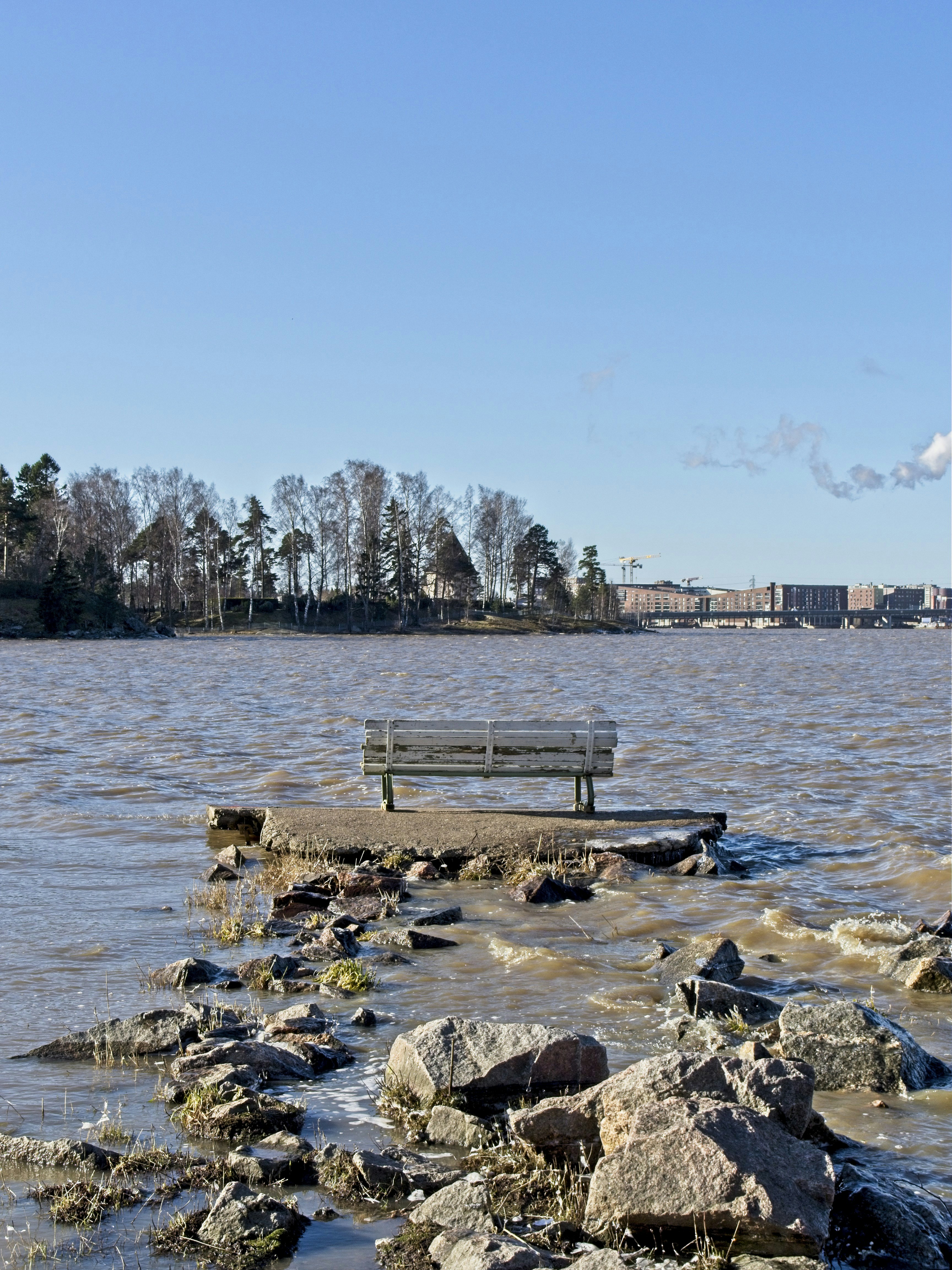 A lone bench sits on a jagged stone jetty extending into a choppy lake under a bright blue sky. Distant buildings on the far shore and a plume of smoke add a touch of industrial scale.