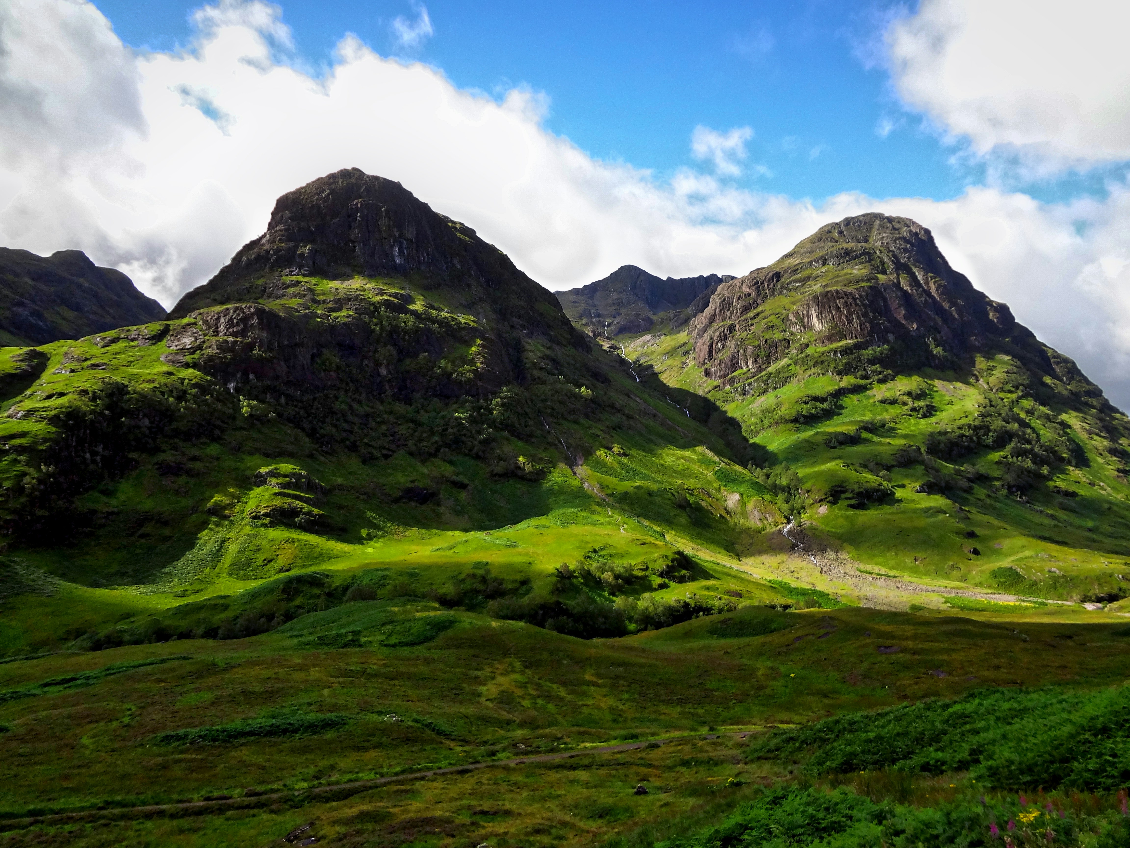 Lush green hills rise majestically under a partly cloudy sky in the Scottish Highlands. The rugged terrain showcases the natural beauty of the landscape.