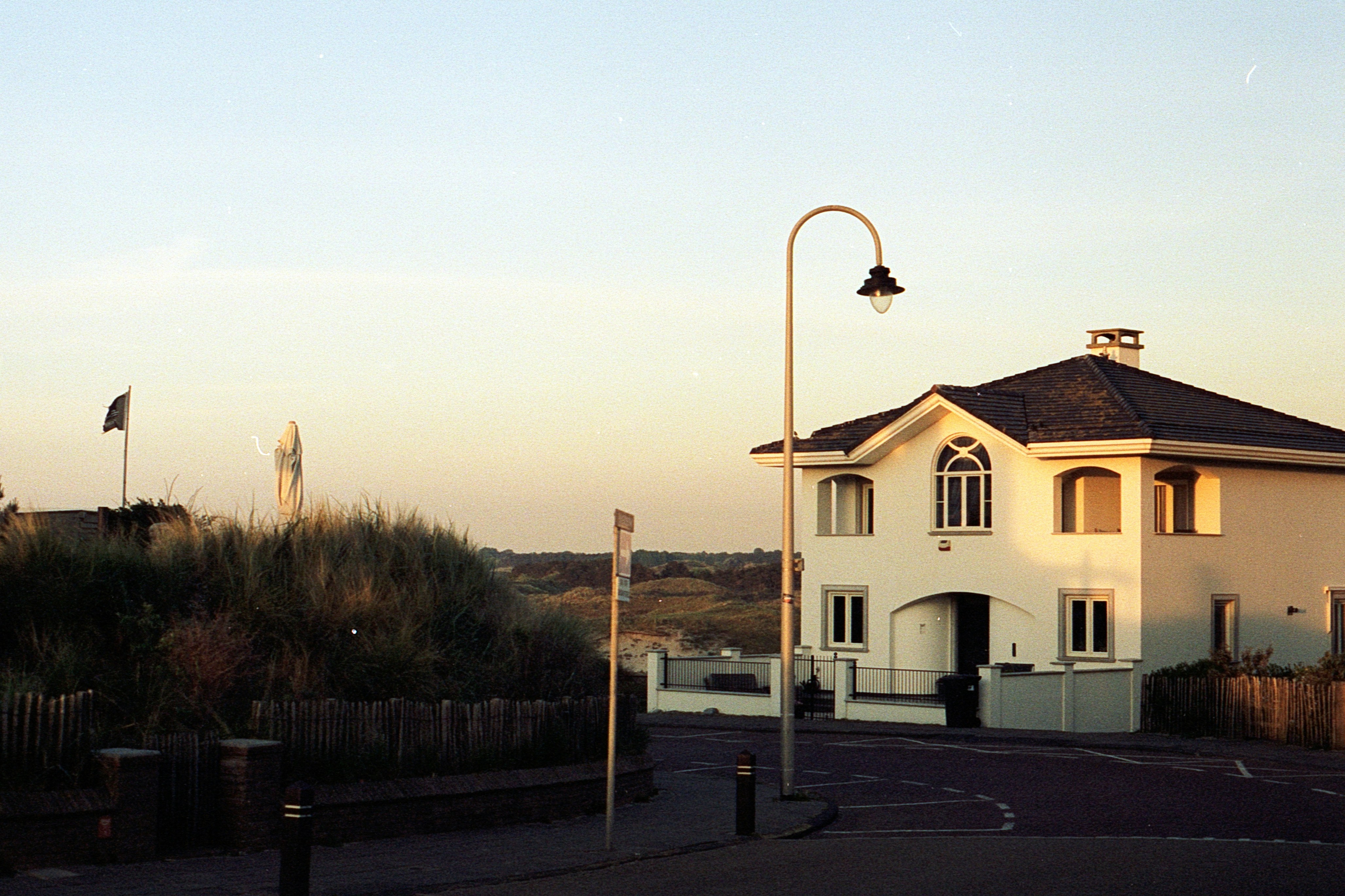Photograph of a coastal dune scene featuring a white villa and a softly glowing streetlamp at sunset, with a quiet road leading to the house.
