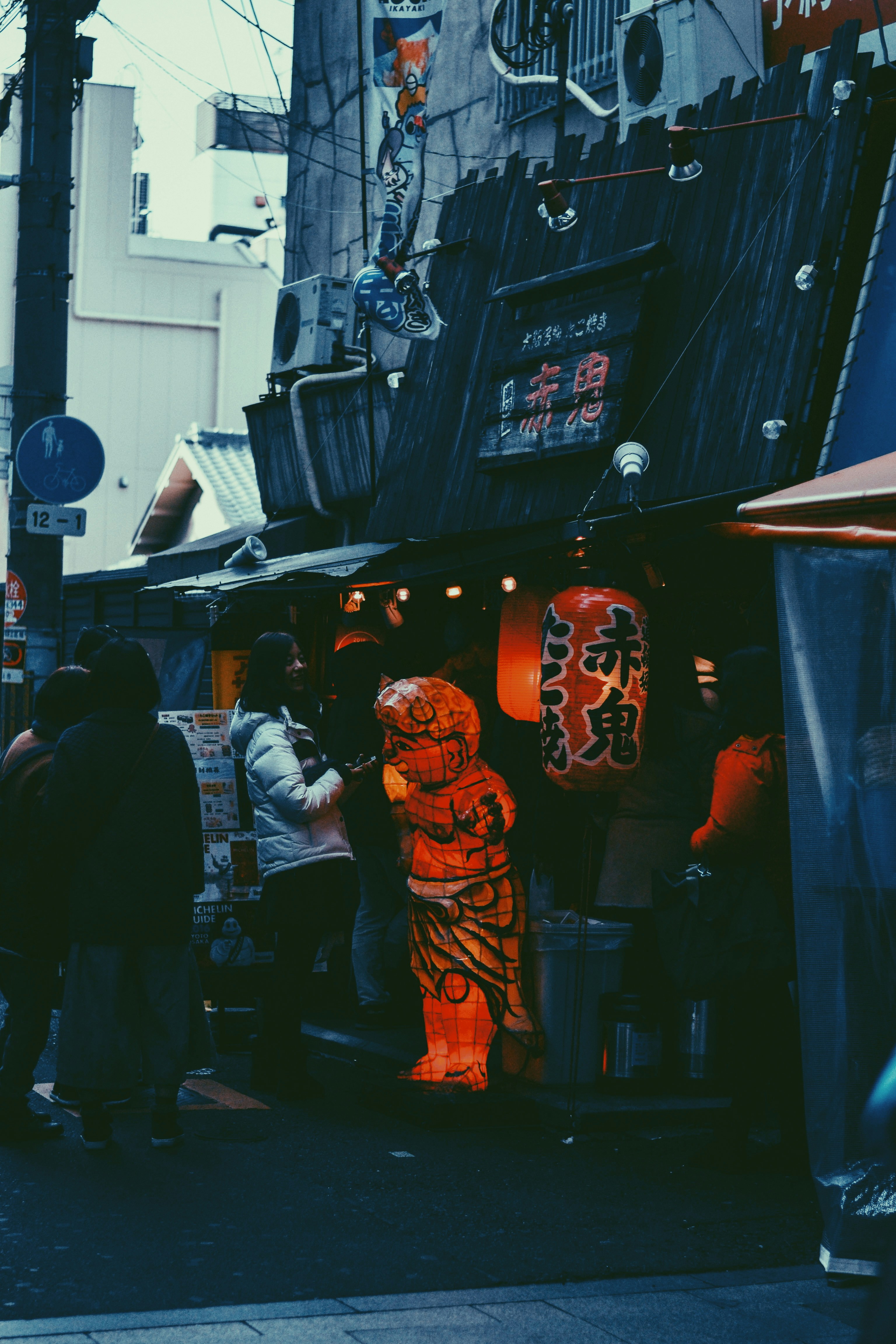 A vibrant lantern figure stands outside a bustling eatery, surrounded by patrons enjoying the lively atmosphere. The rich colors and urban setting highlight local culture.