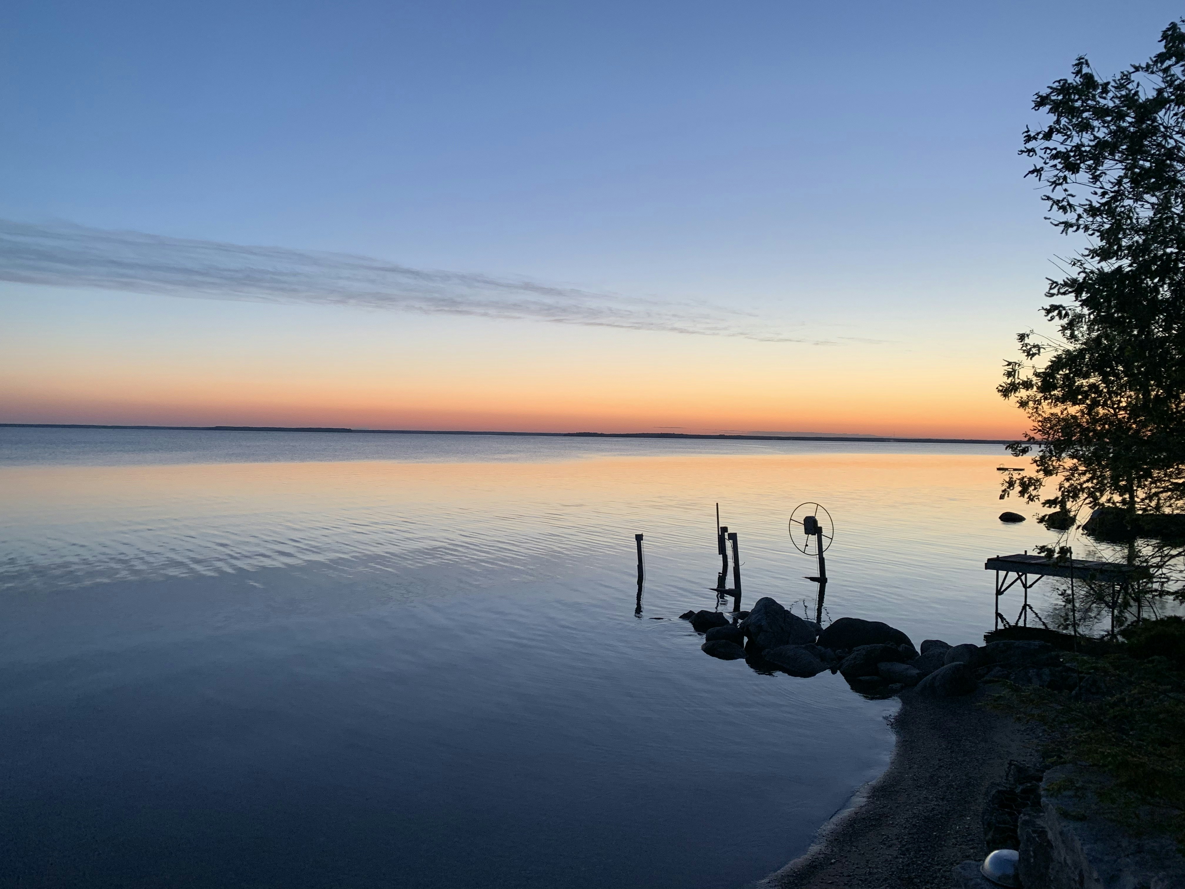 Tranquil waters meet the horizon at dusk, framed by silhouettes of rocks and a tree under a gradient sky.