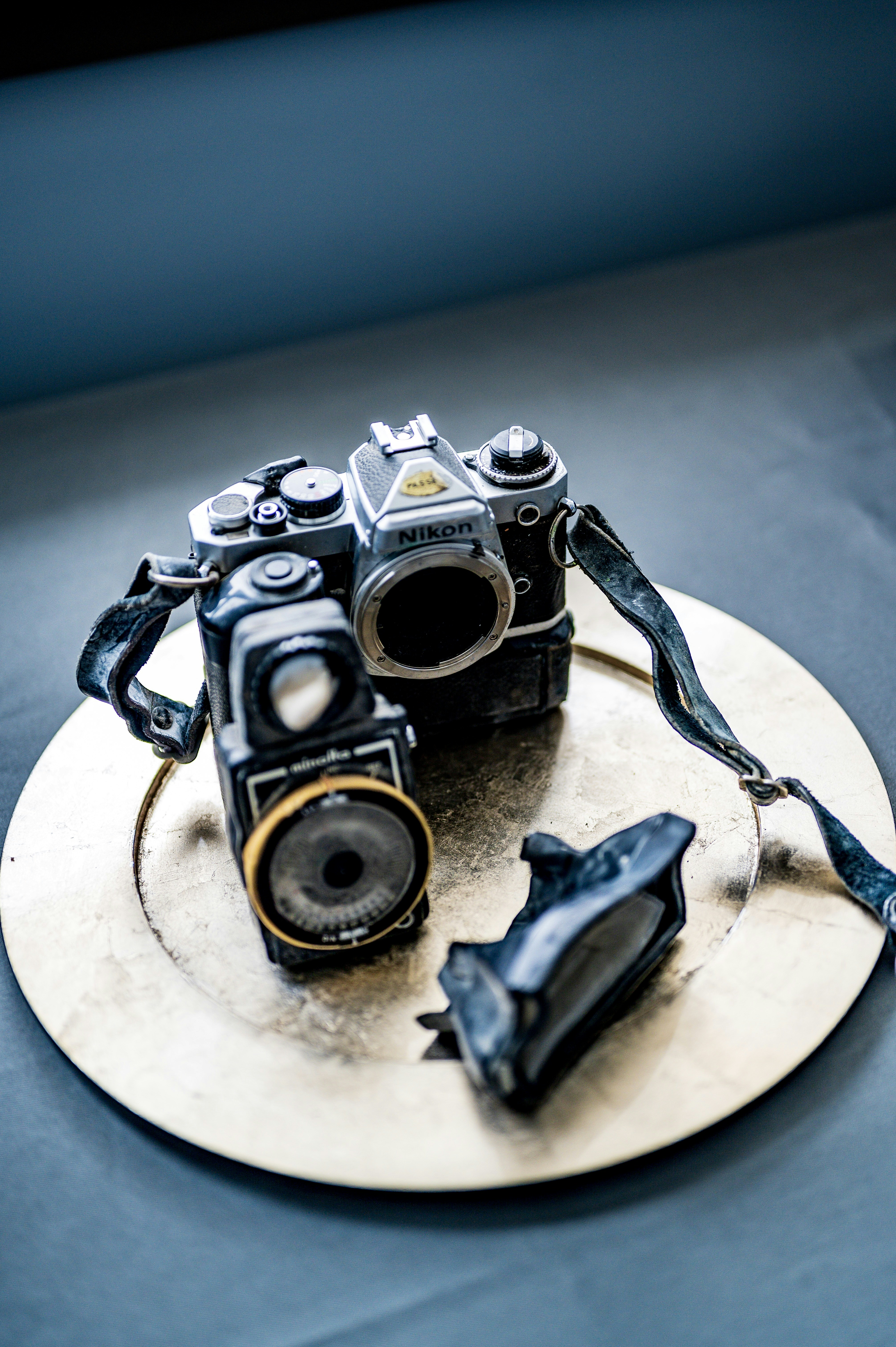 Pair of vintage Nikon cameras placed on a circular stand with a soft, moody backdrop; the composition highlights aged lenses and worn straps. This is a still-life photograph capturing a collector's relic.