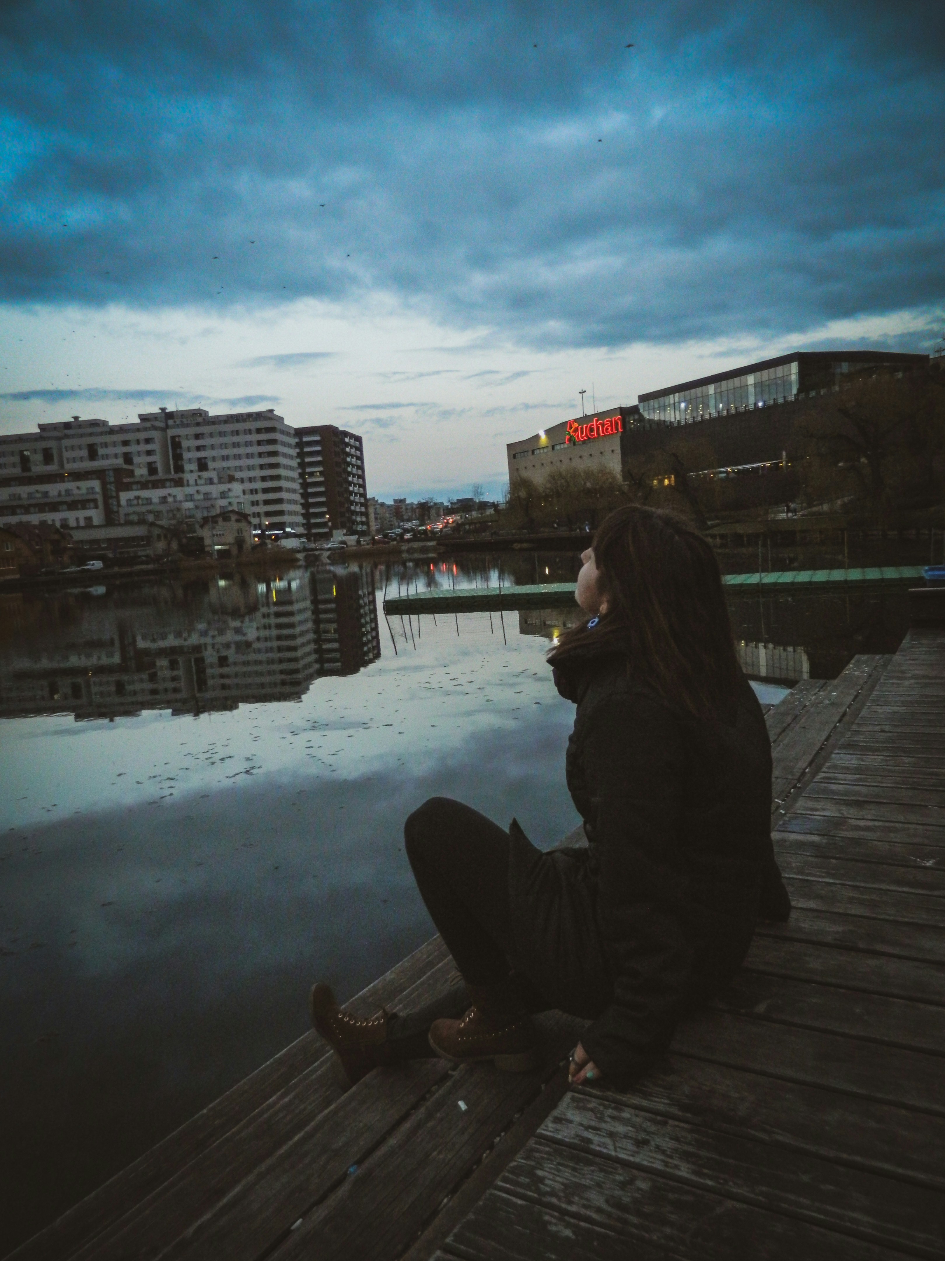 A contemplative figure sits on a wooden dock, gazing at the water's surface reflecting the city skyline under a twilight sky.