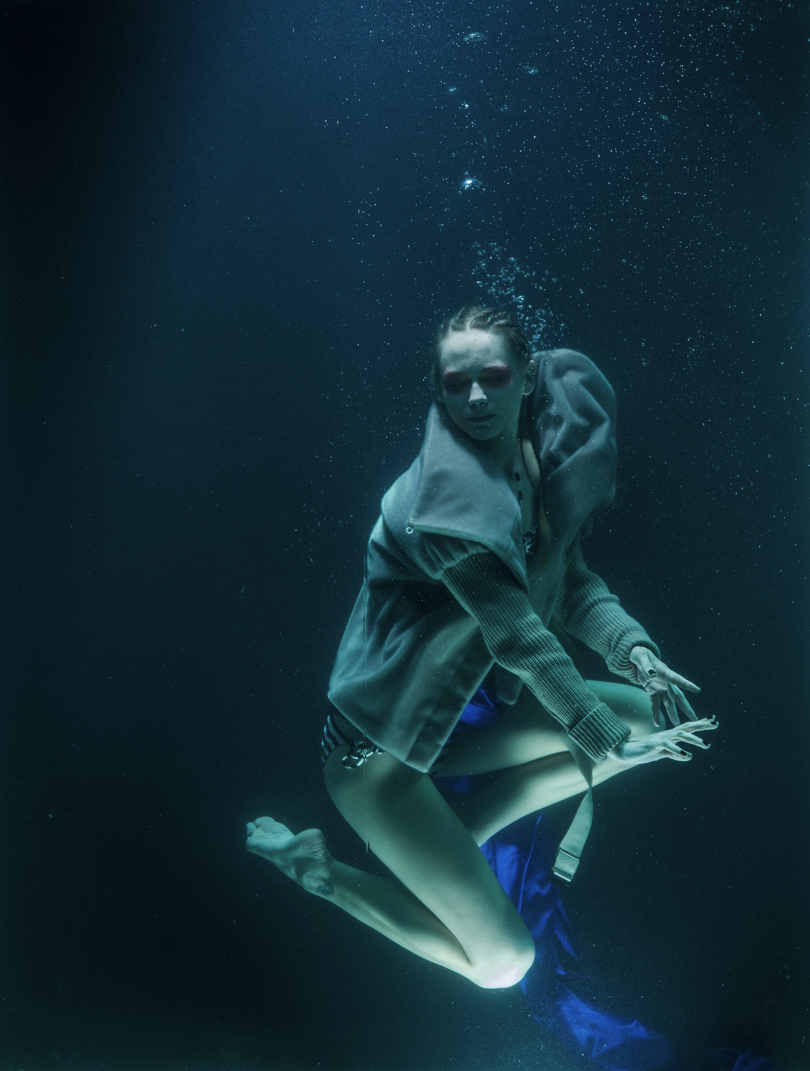 Underwater portrait of a model in a gray coat and blue bikini, suspended in dark blue water with bubbles, a moody photograph.