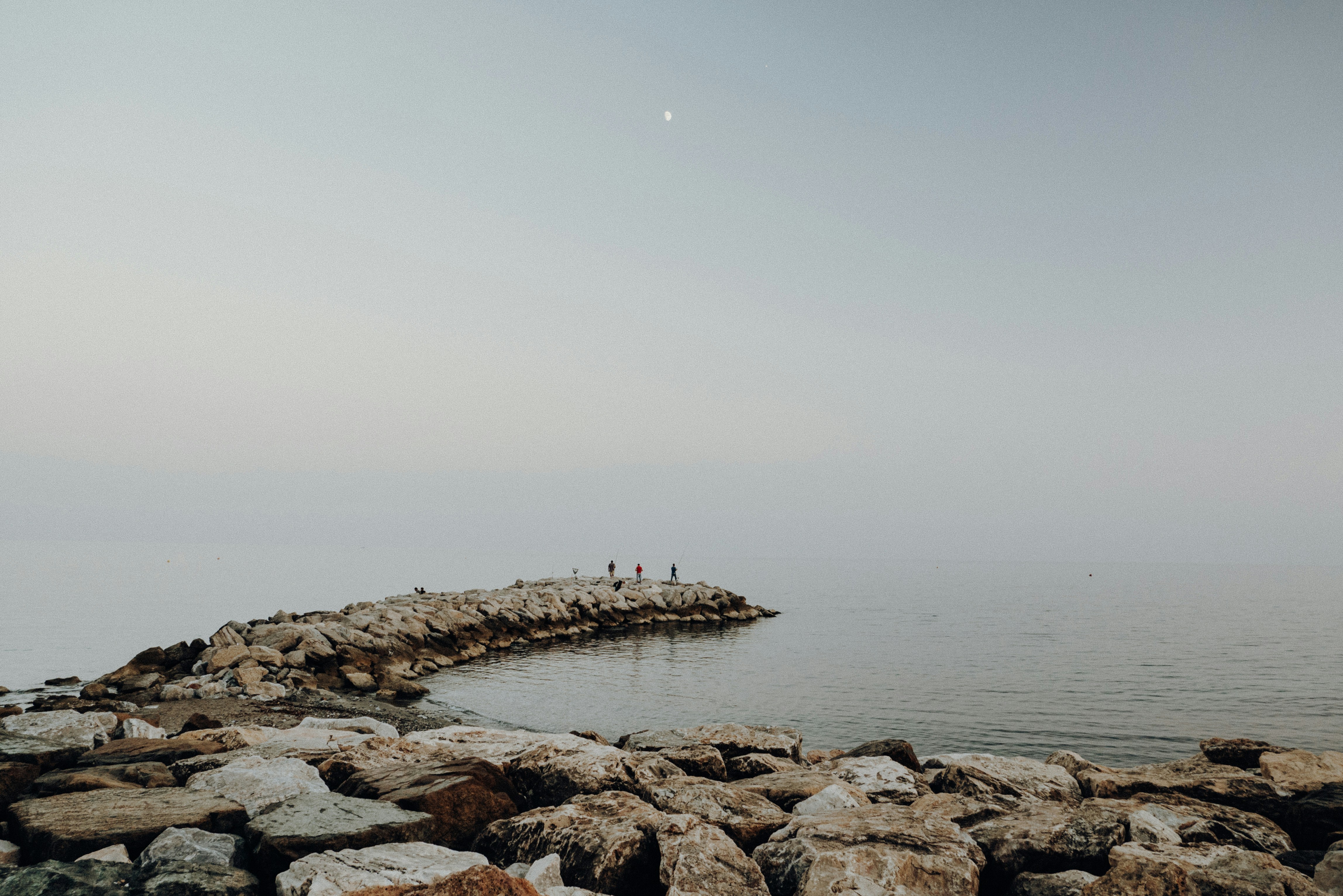 People walking along a rock jetty extending into a calm sea under a pale sky.