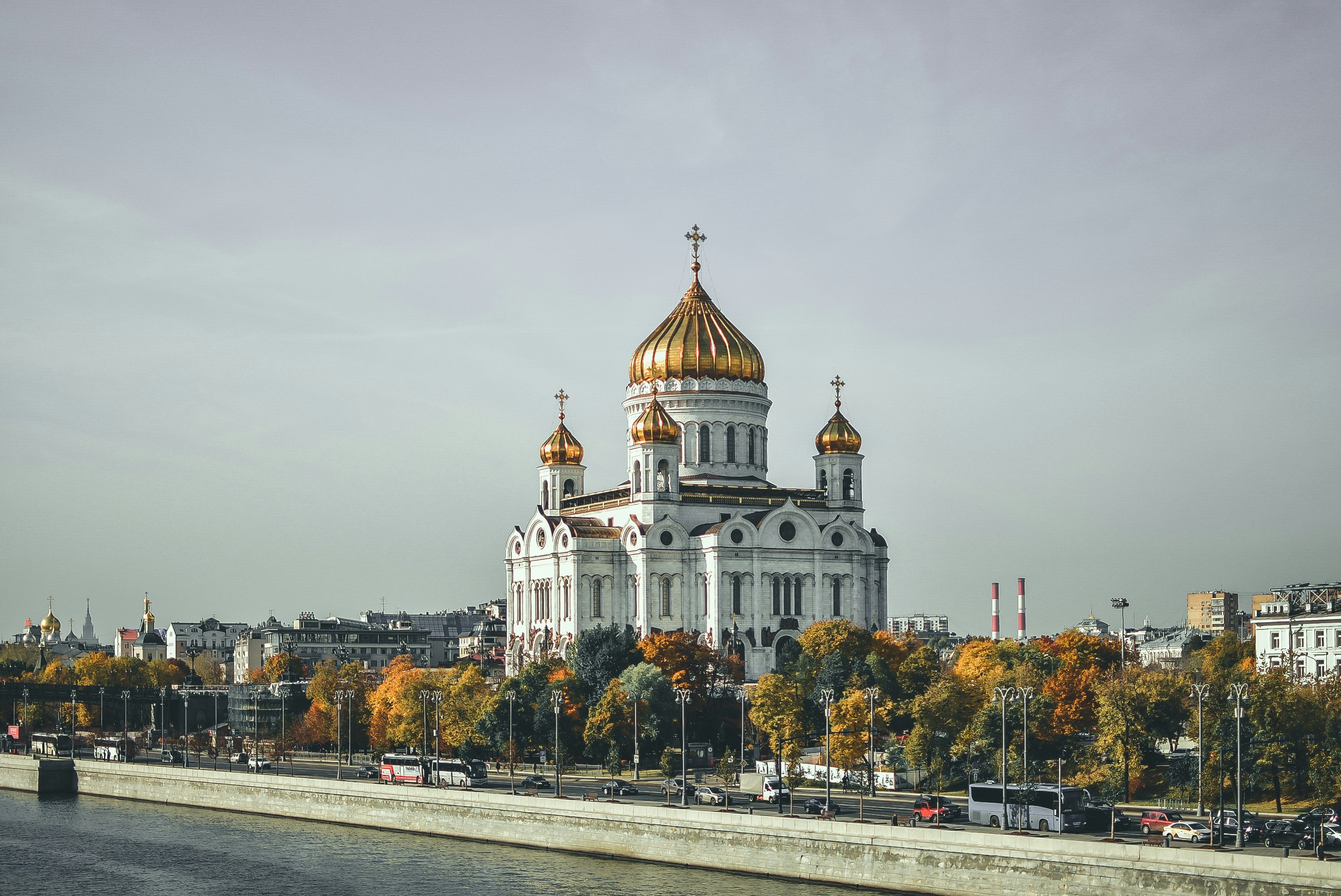 Ornate cathedral with golden domes surrounded by vibrant autumn foliage along a riverbank.