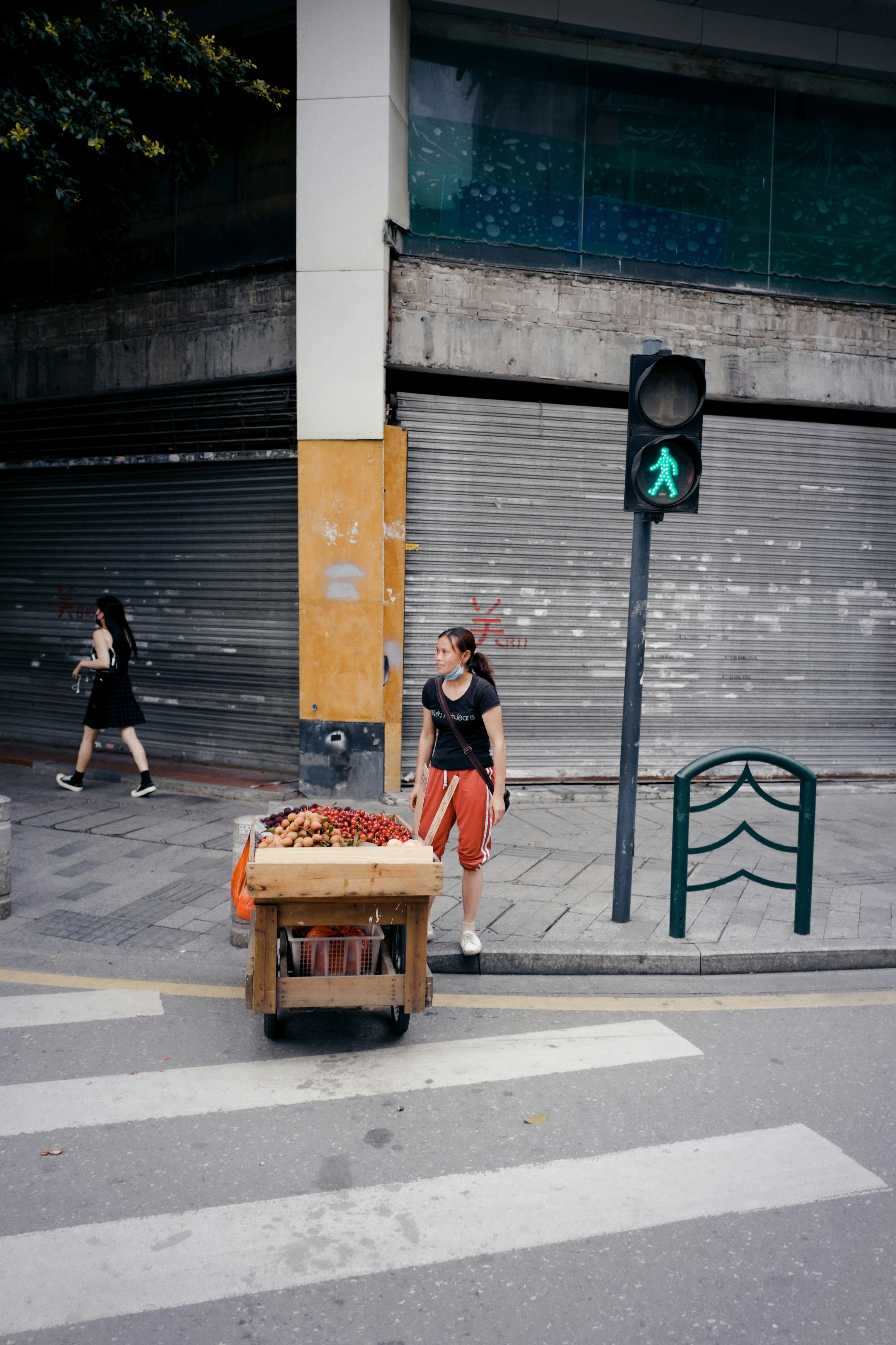 A street vendor with a cart of fruits stands at a crosswalk, illuminated by a green pedestrian signal, while a passerby walks in the background.